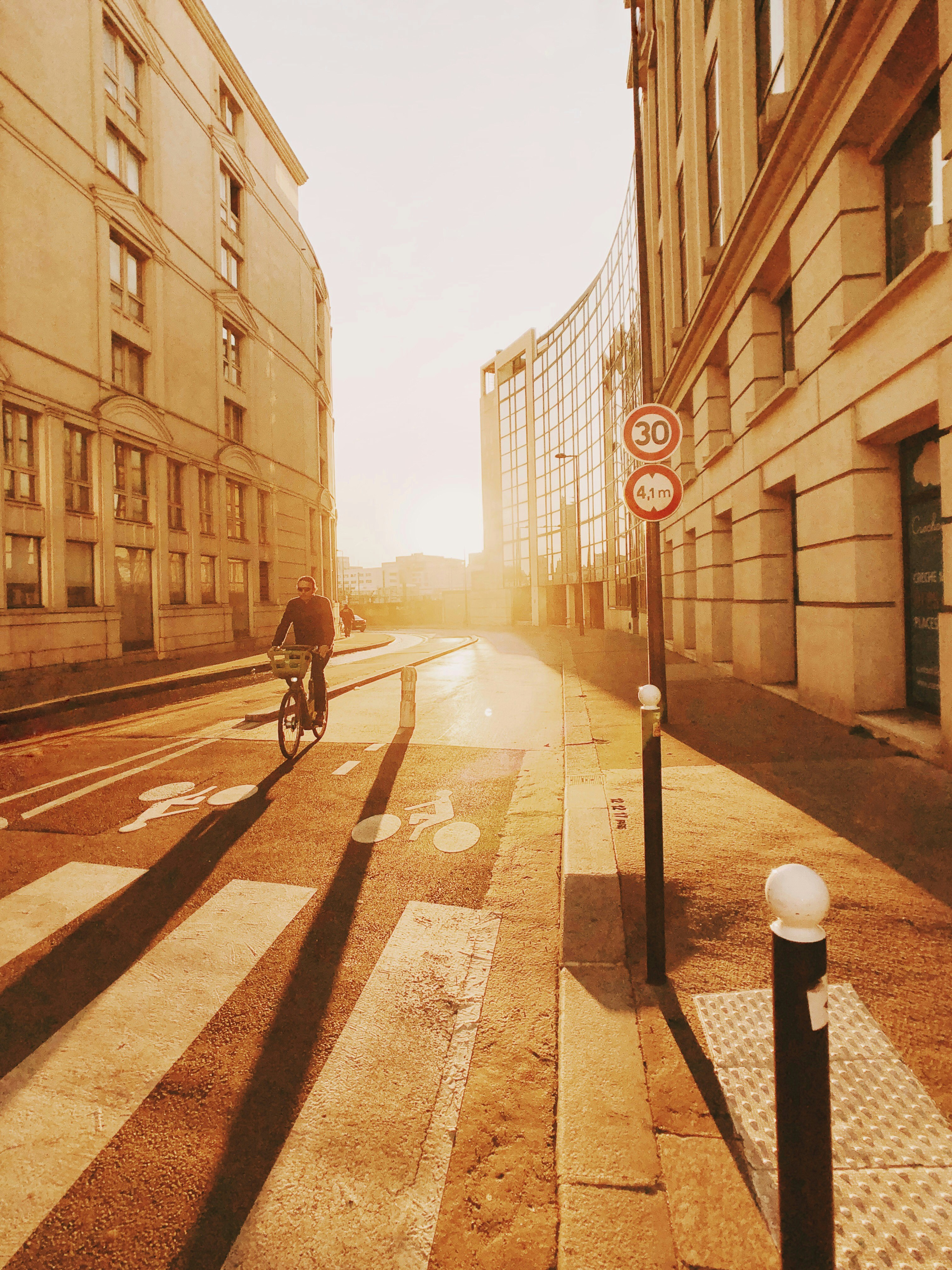 A cyclist rides along a sunlit street, flanked by modern and classic architecture, while long shadows stretch across the pavement.