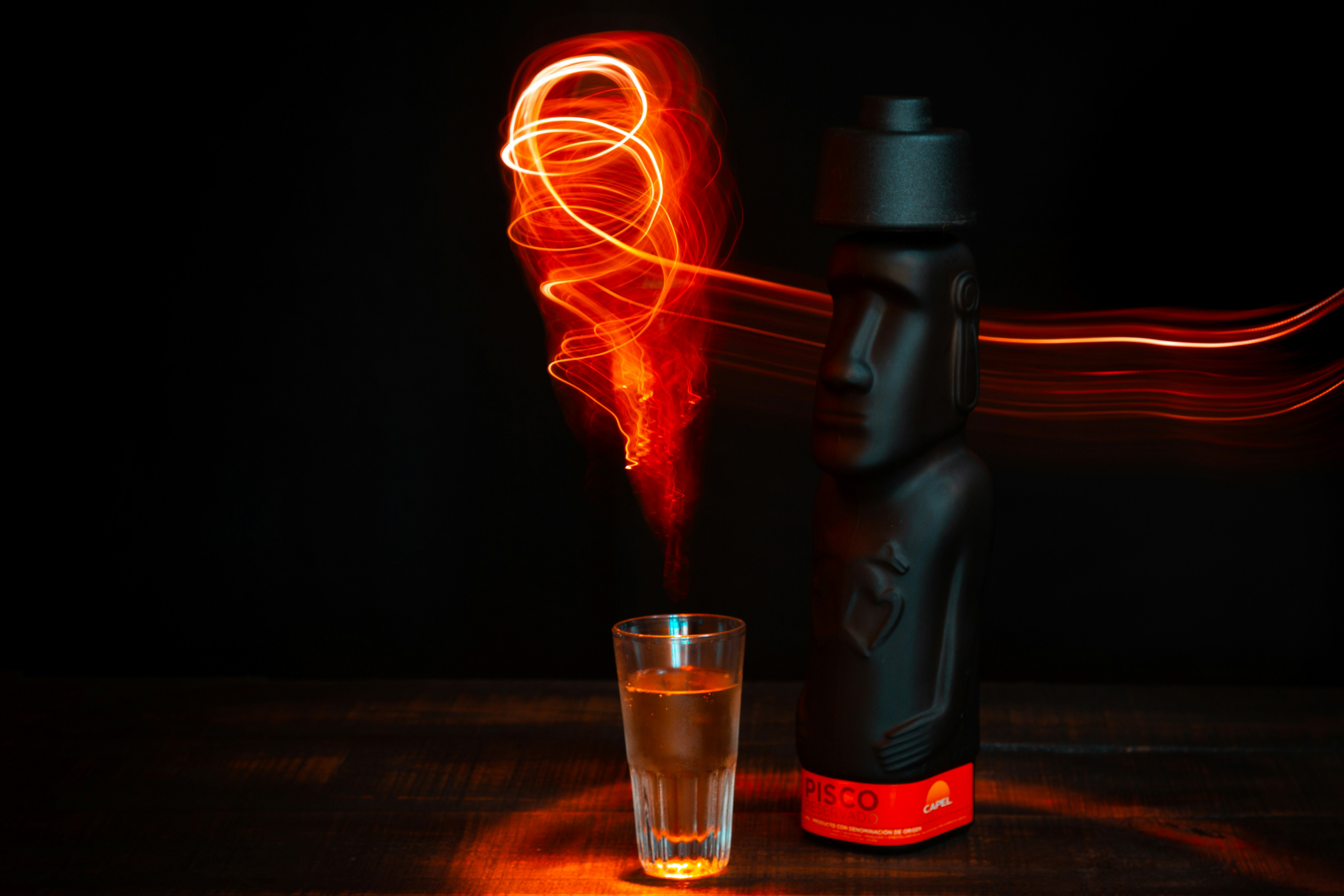 A dark, artistic composition featuring a black bottle resembling a Moai statue beside a shot glass, illuminated by swirling light trails.