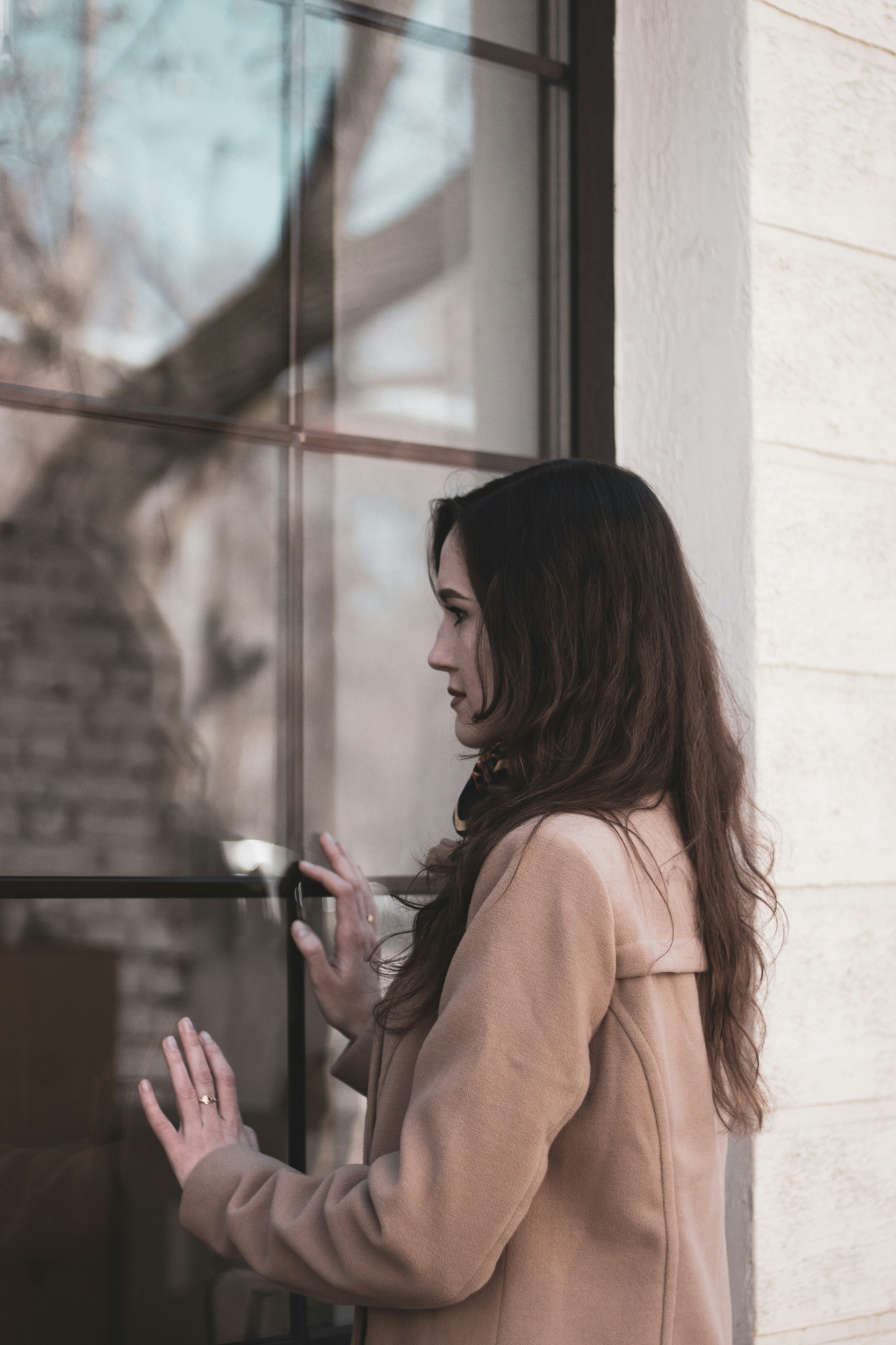 Woman in a beige coat gazes thoughtfully at a window, with soft reflections of nature in the glass.