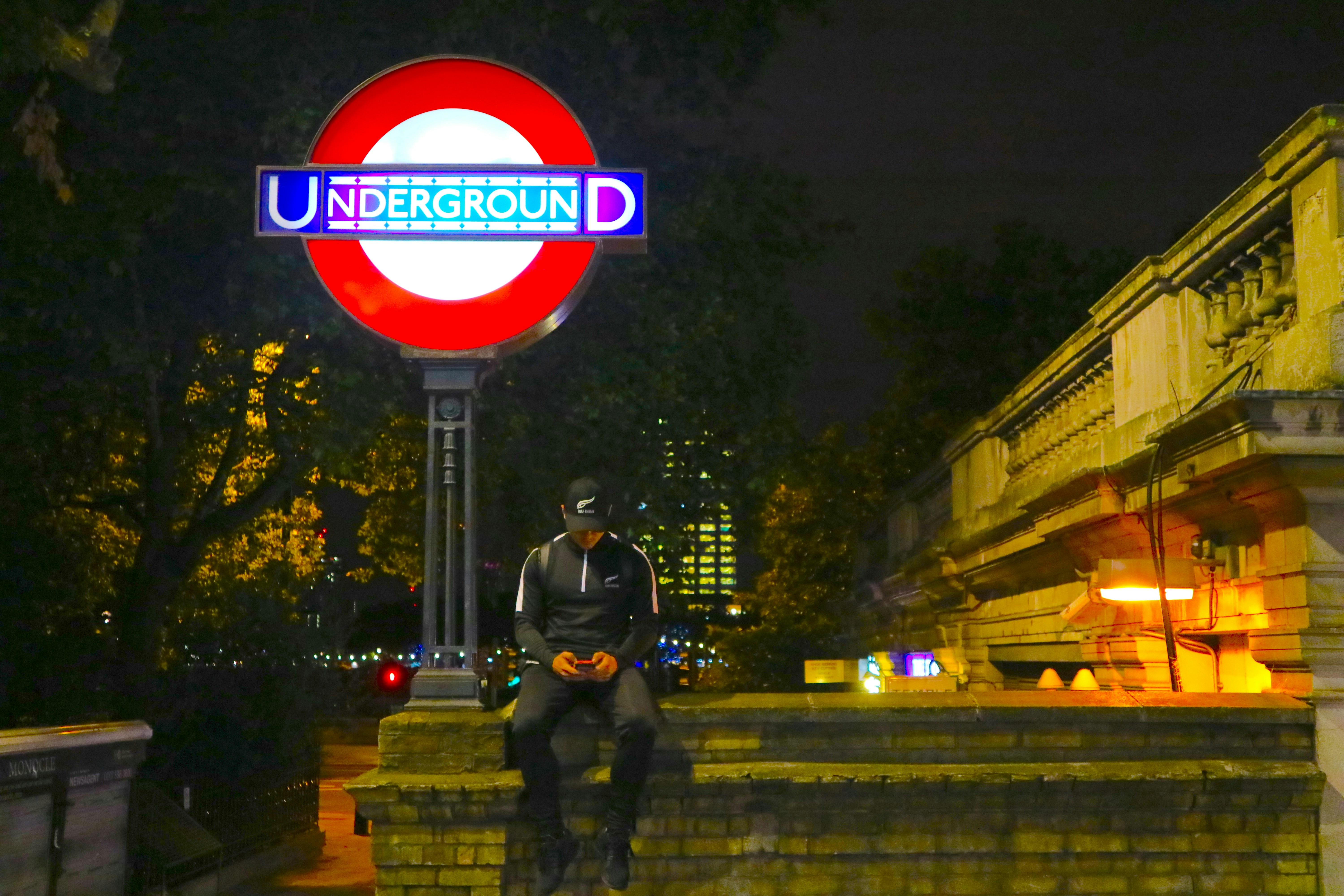 Nighttime street photograph capturing a lone figure seated on a stone ledge beside a lit London Underground sign. The composition emphasizes quiet urban atmosphere and solitary pause.