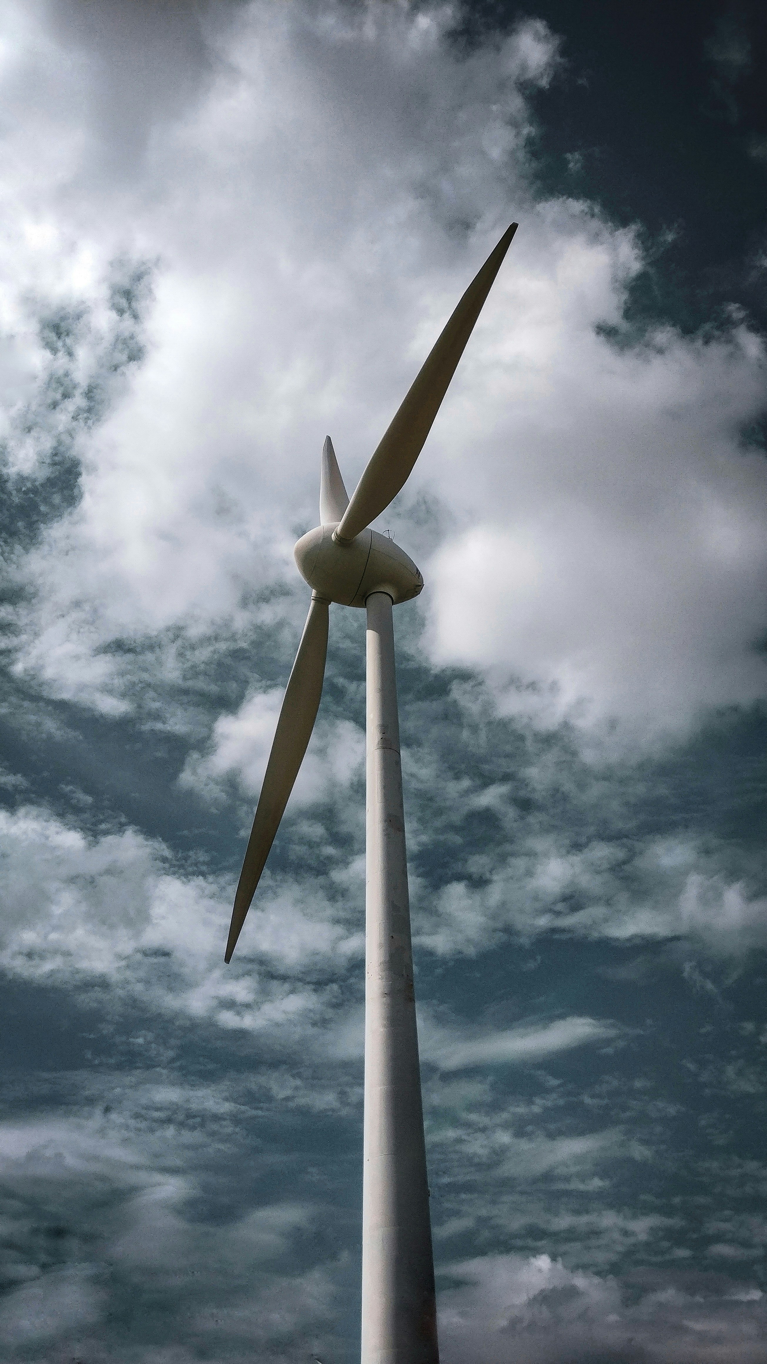 Tall wind turbine rises into a partly cloudy sky, blades framed against blue and white clouds. This photo emphasizes scale and the interaction between structure and weather.