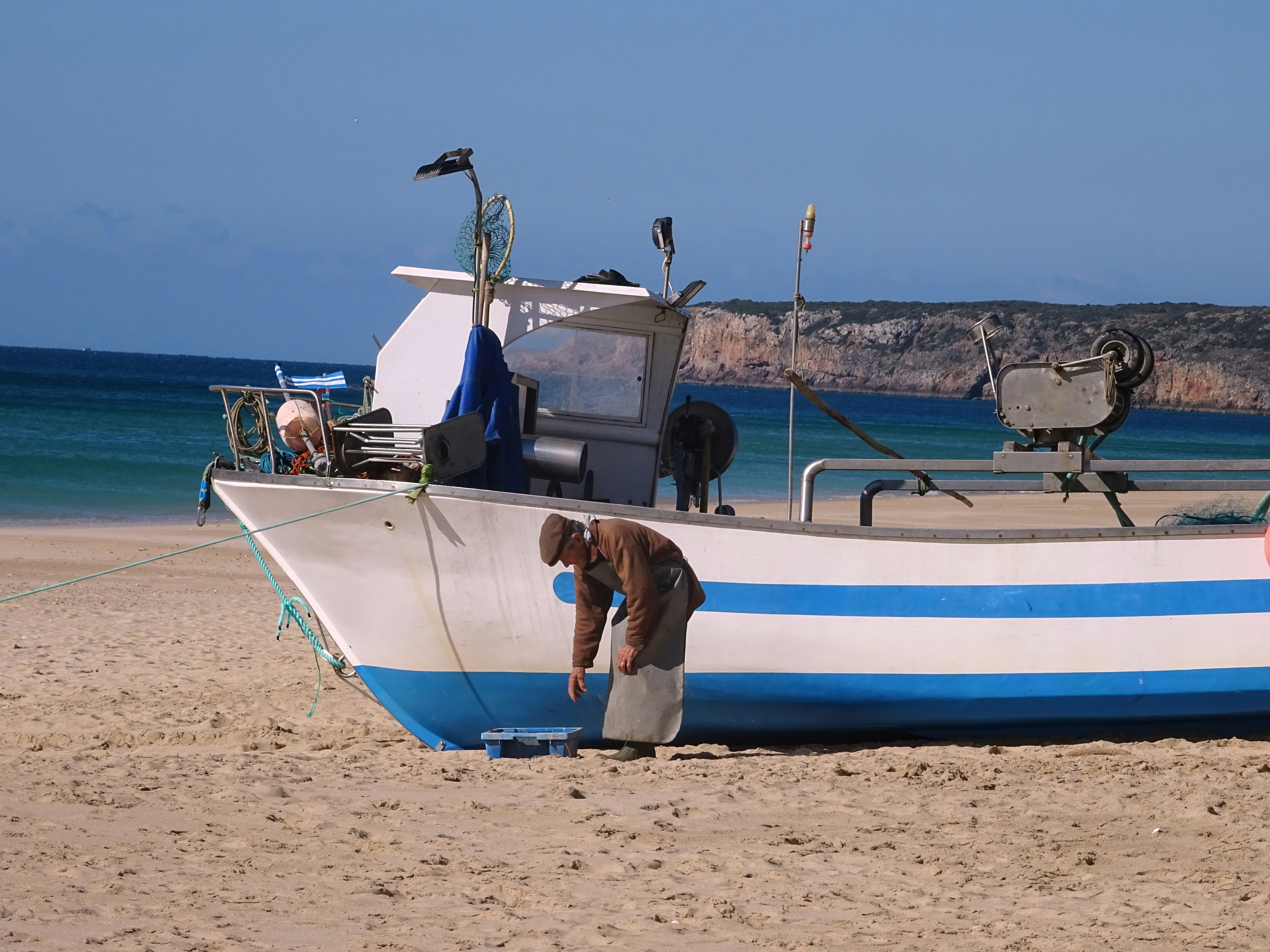 Fisherman inspecting gear on a traditional boat along a serene beach, with gentle waves lapping at the shore.