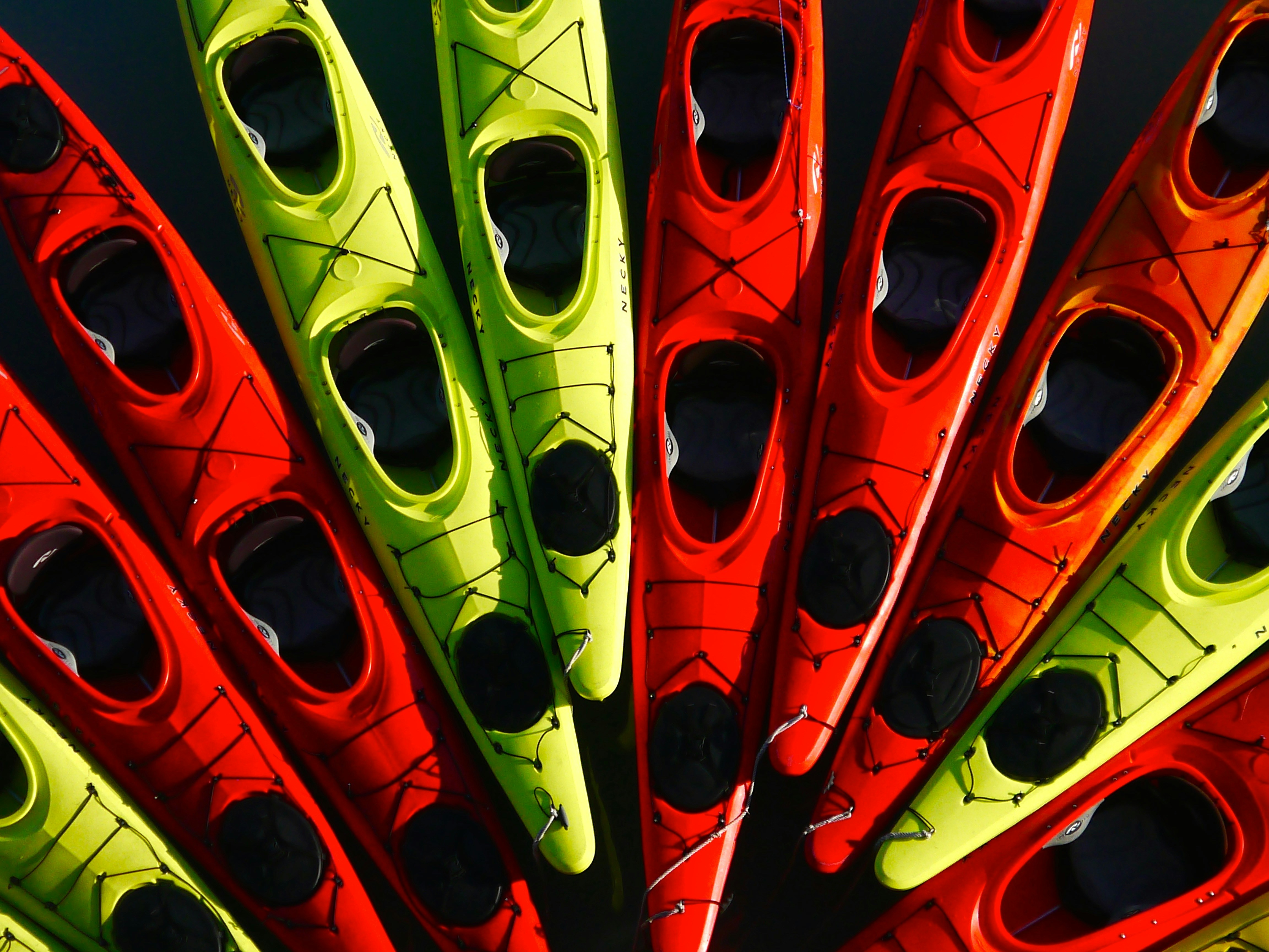 Top-down view of neon red, lime green, and orange kayaks arranged in a circular fan, forming a bold geometric pattern.