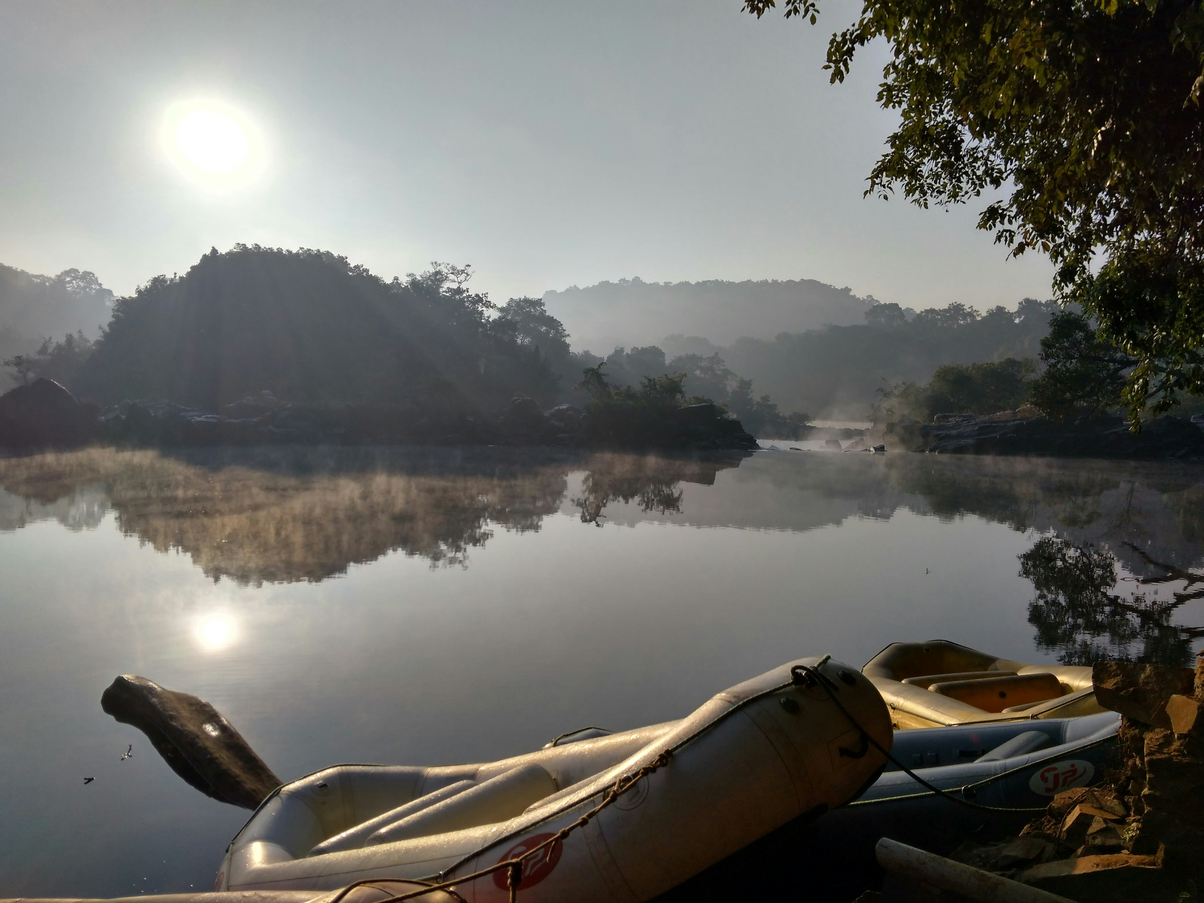 Calm river scene at dawn, featuring boats resting on the shore with mist rising from the water and sun illuminating the landscape.
