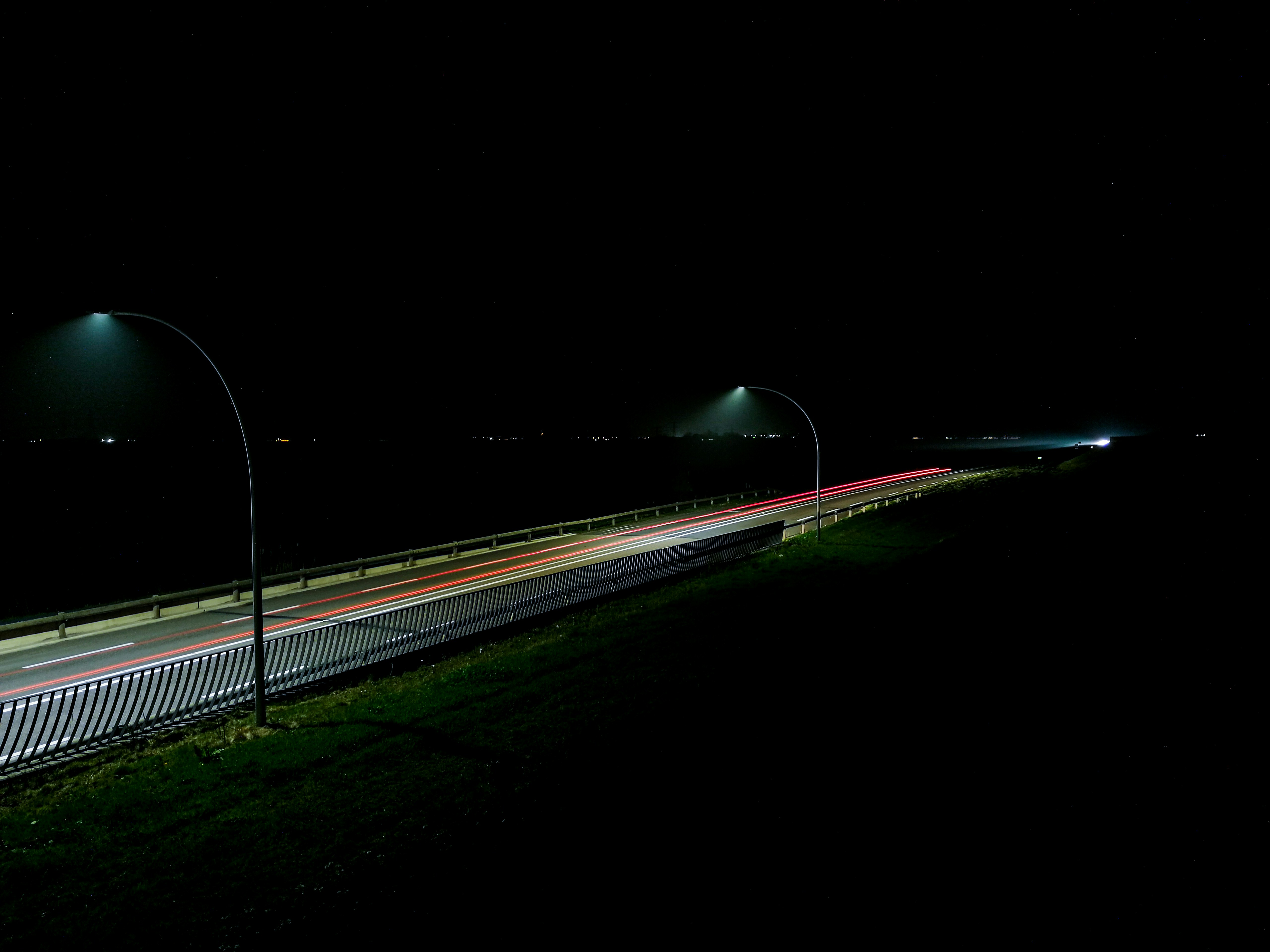 Long exposure shot capturing light trails from vehicles on a dimly lit road, framed by streetlights. The darkness enhances the vibrant colors of the moving lights.