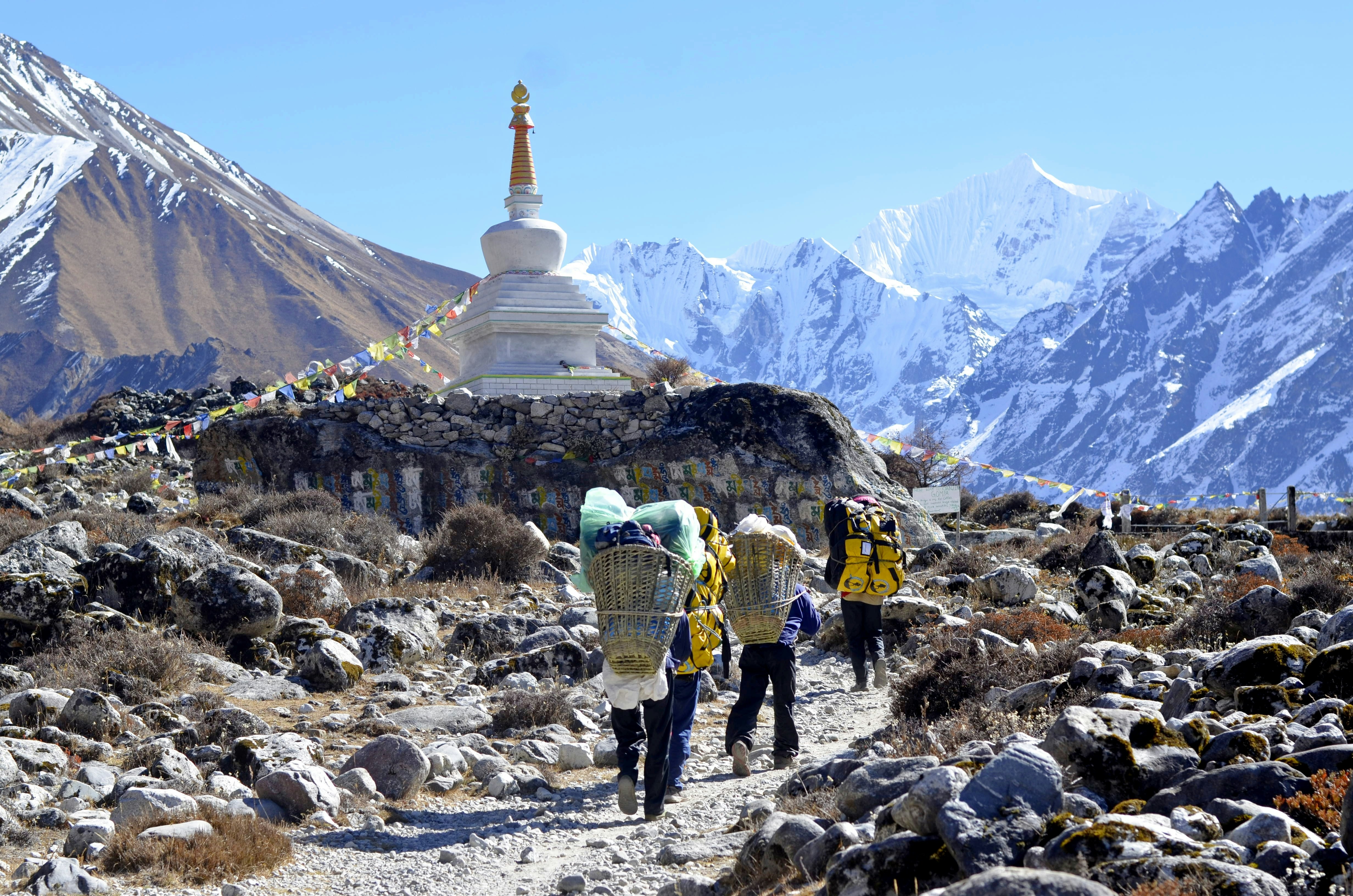 Porters navigate a rocky path towards a Buddhist stupa, framed by majestic snow-capped mountains and colorful prayer flags.