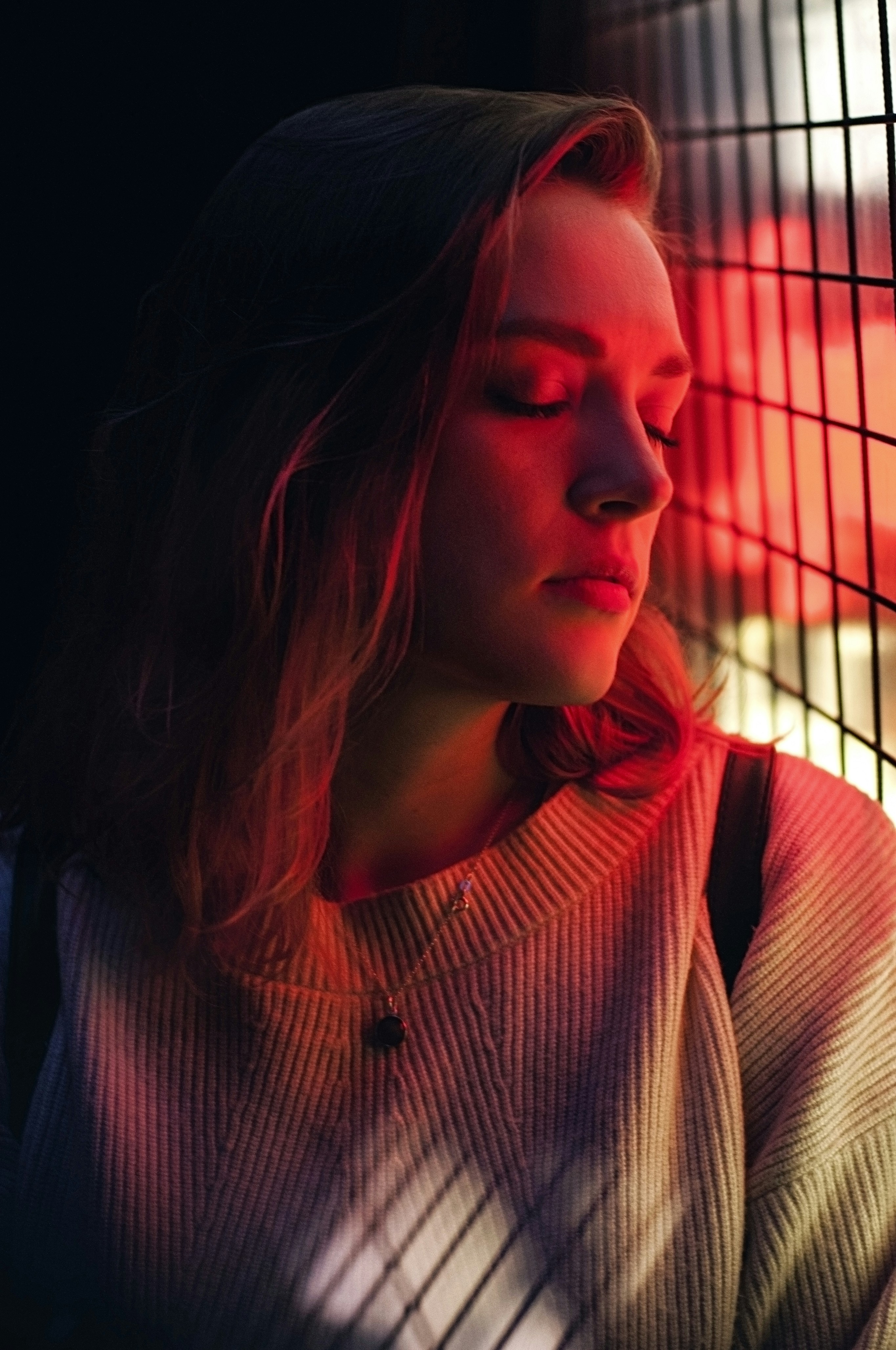 A contemplative woman with closed eyes is illuminated by soft, colorful light against a patterned backdrop of a grid. The play of light creates a serene atmosphere.