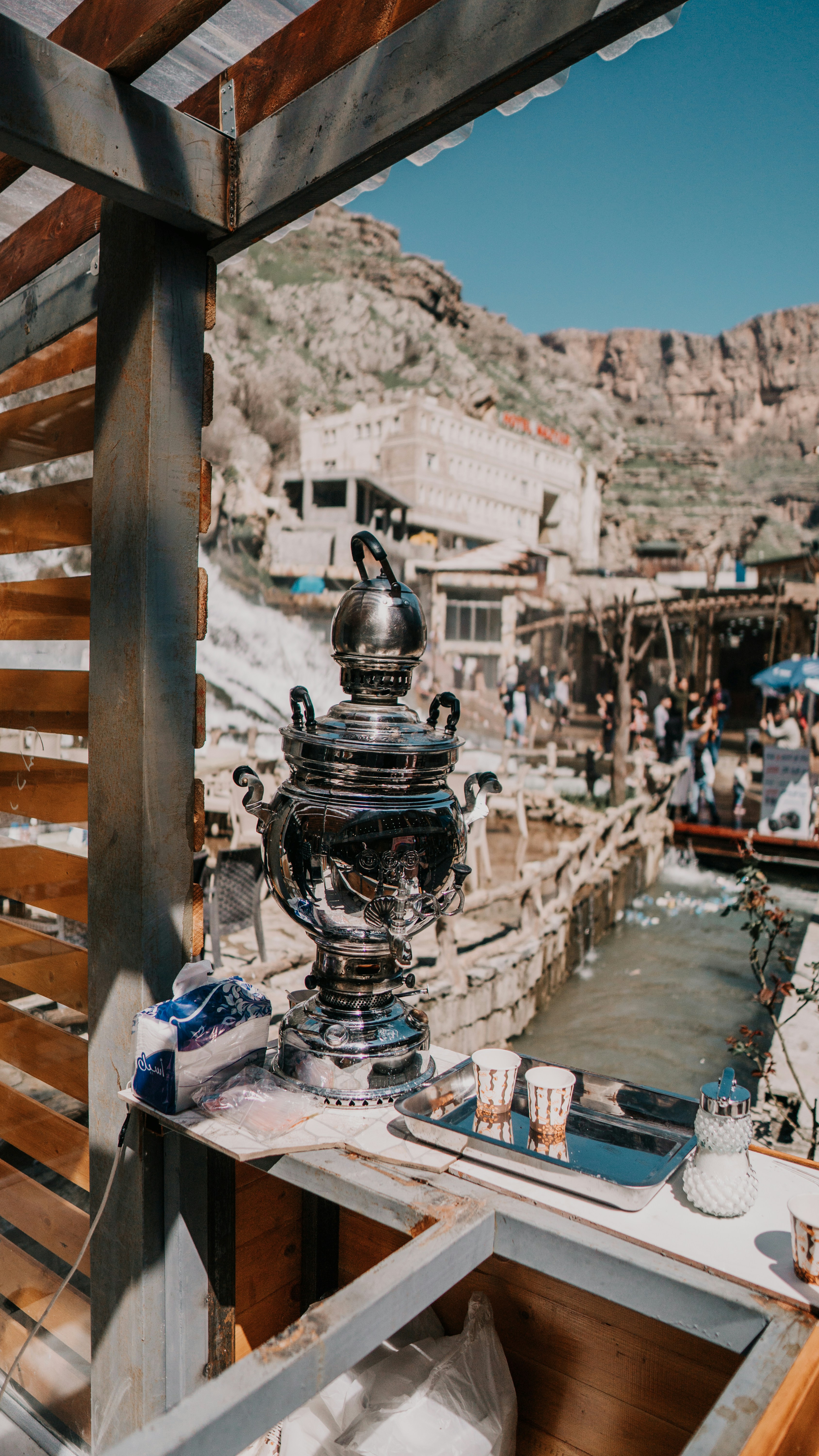 A polished samovar stands prominently on a wooden counter, overlooking a bustling riverside scene with visitors and scenic mountains in the background.