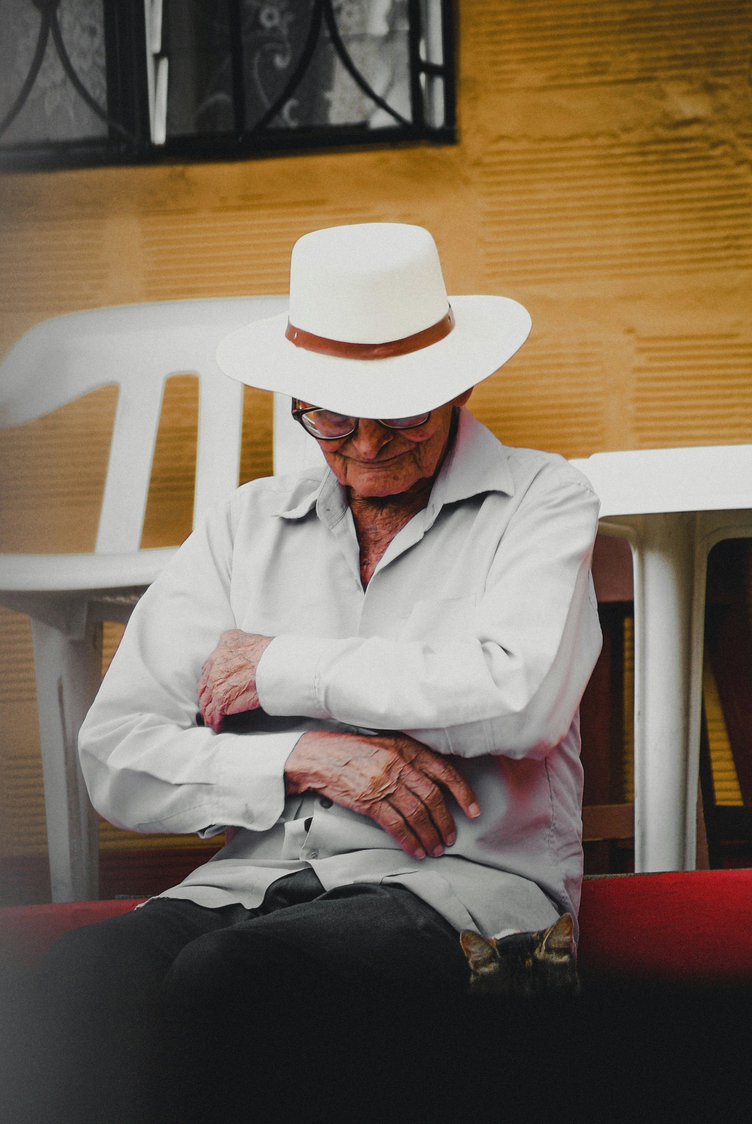 Elderly man in a white hat and shirt sits thoughtfully, arms crossed, in a warm-toned environment. A cat rests nearby, adding a touch of tranquility.