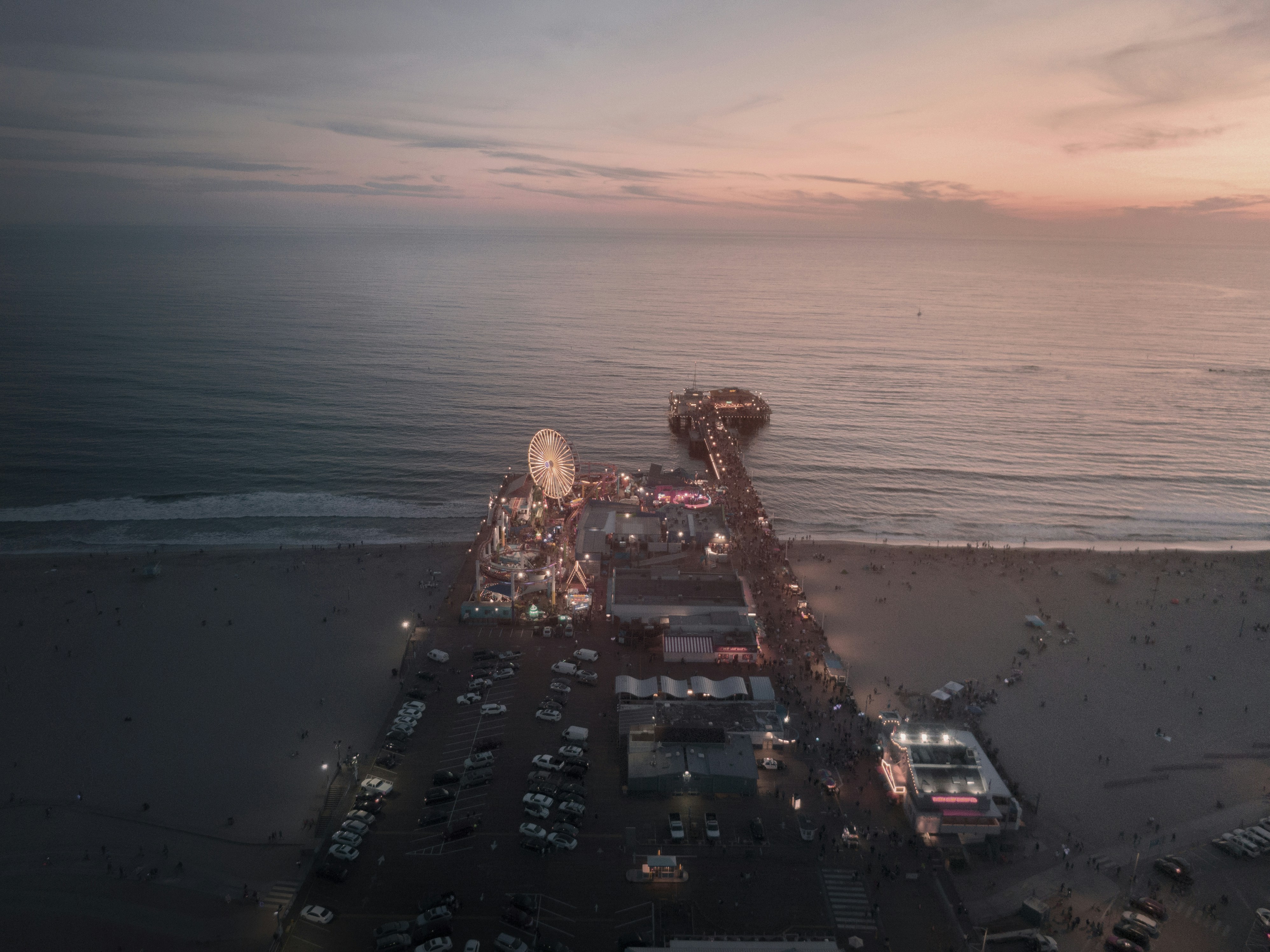 Aerial drone photograph of a seaside pier at dusk, featuring a lit Ferris wheel and glowing boardwalk extending into the ocean.