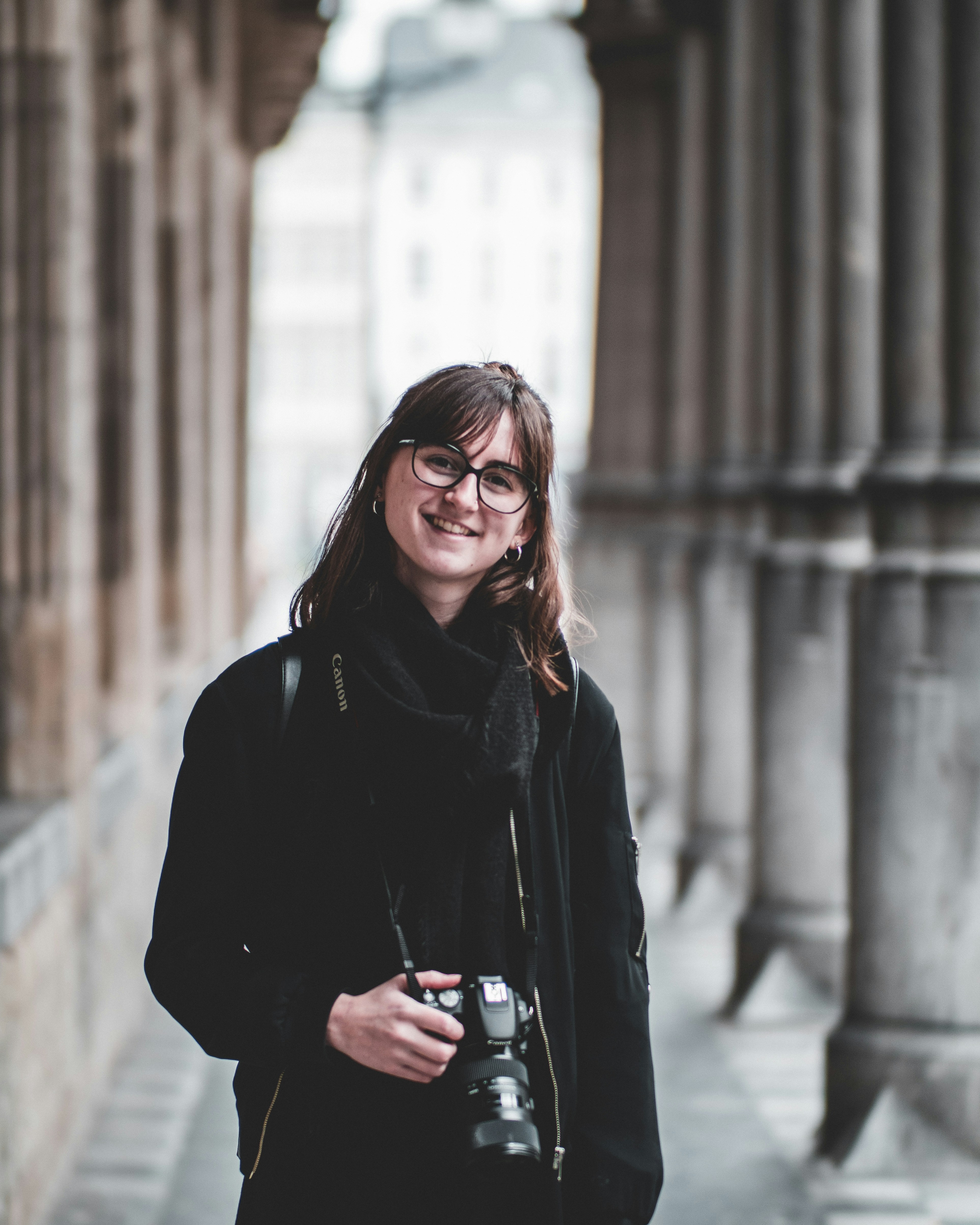 A photographer stands confidently in an archway, holding a camera, with historical columns framing the scene. The soft light highlights the subject's joyful expression.