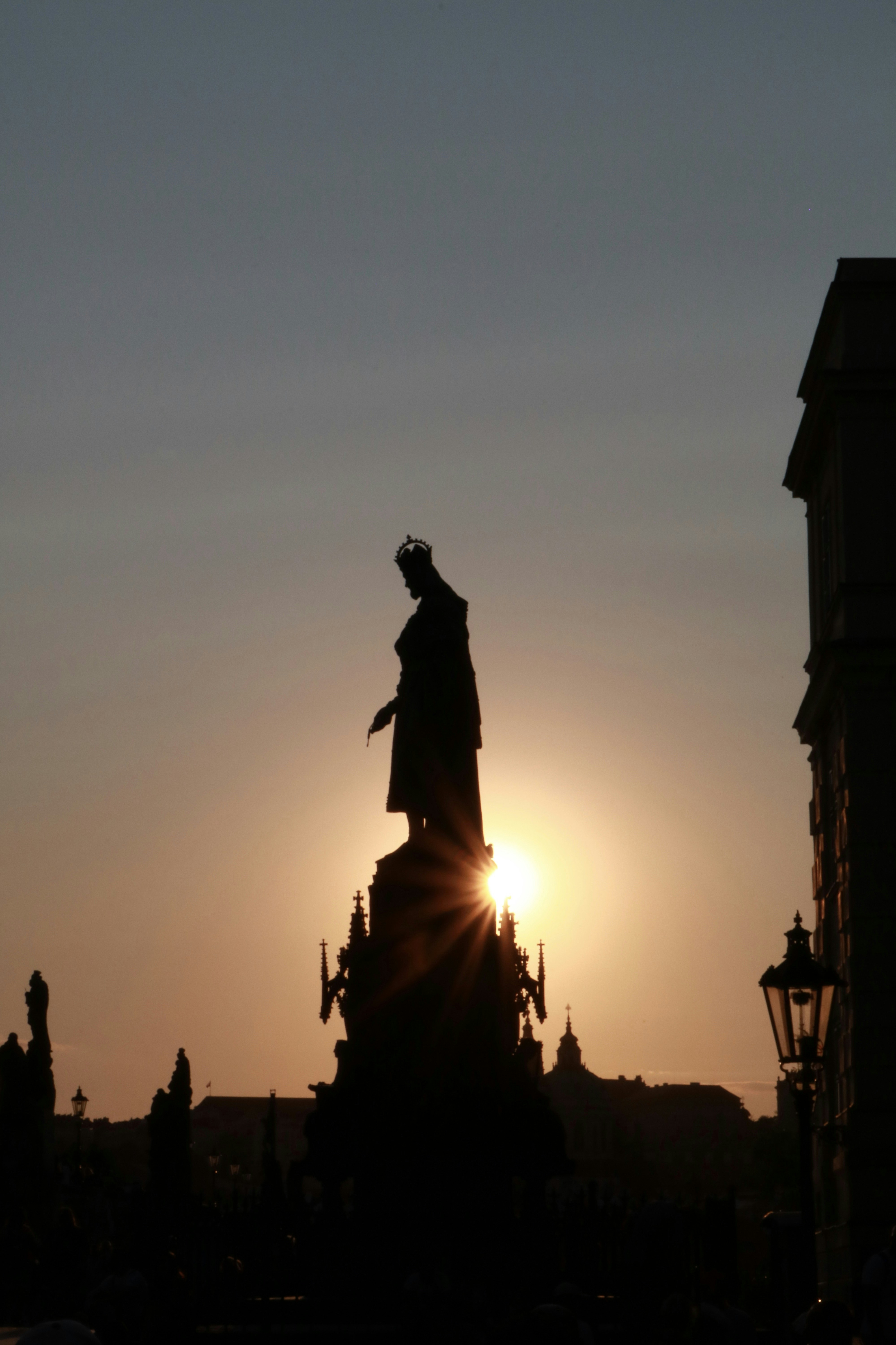 Silhouette of a statue against a glowing sunset, framed by ornate streetlamps and distant architecture.