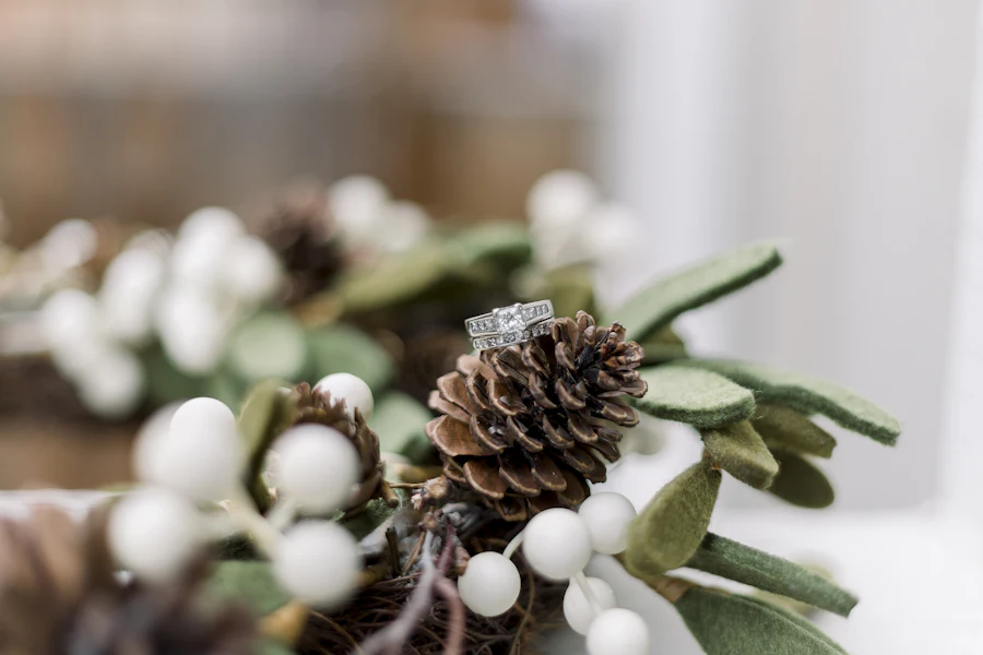 Close-up of a holiday wreath with pinecones, white berries, and green felt leaves, softly blurred in the background. A ring catches the light on the central pinecone.