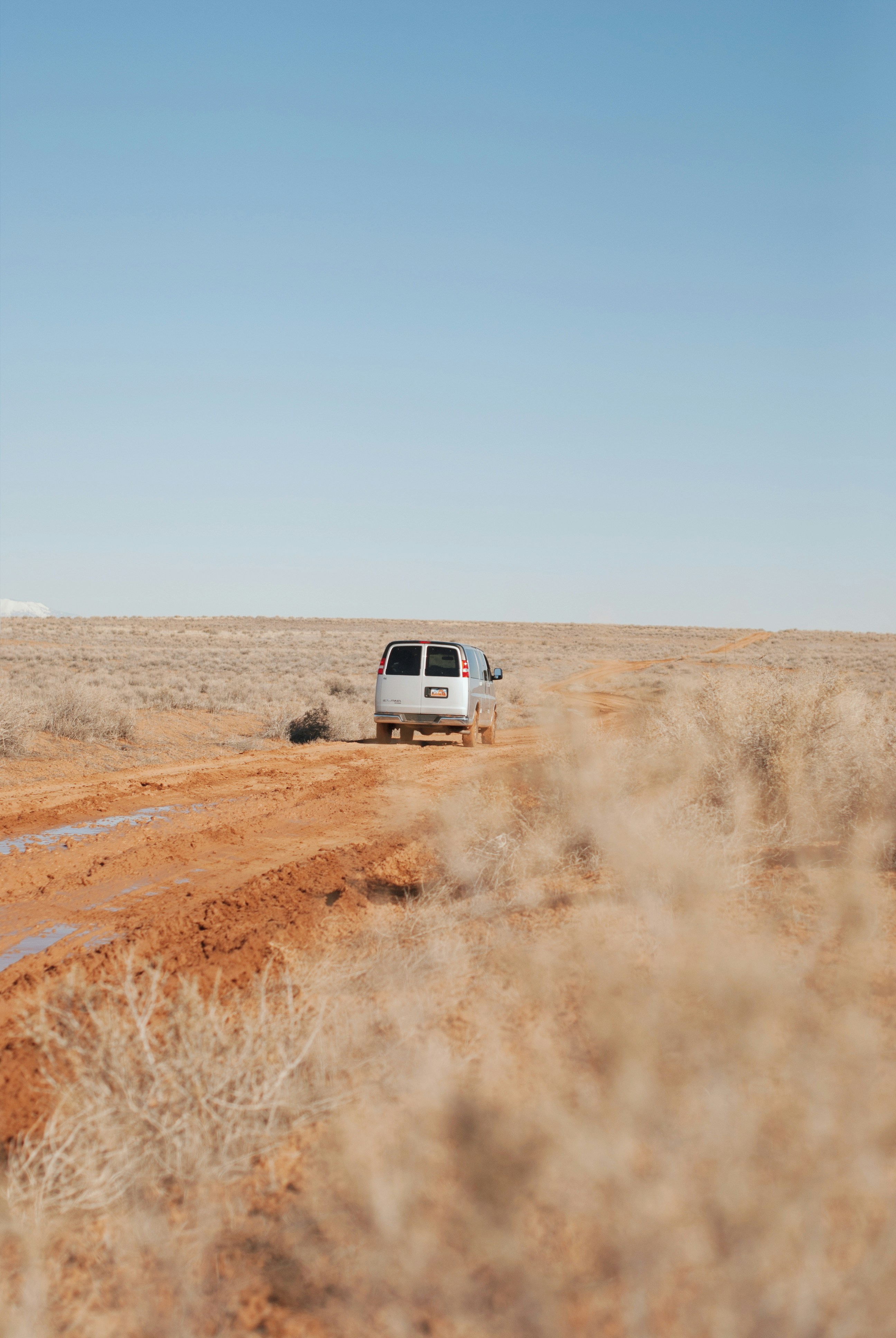 A silver van navigates a dusty road in a vast, dry landscape under a clear blue sky.