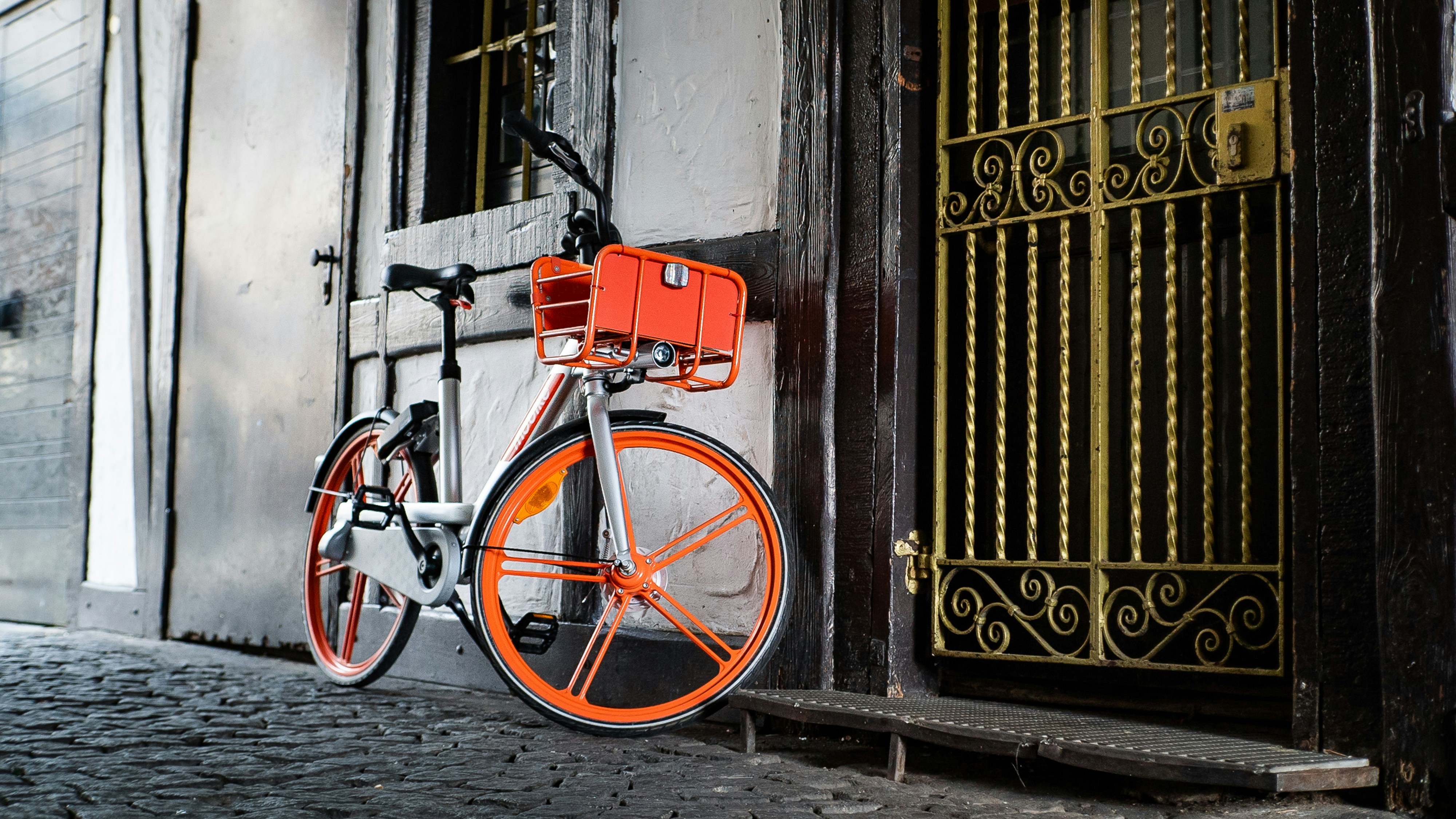 Orange bicycle with a front basket parked beside an ornate wrought iron gate on a cobblestone street.