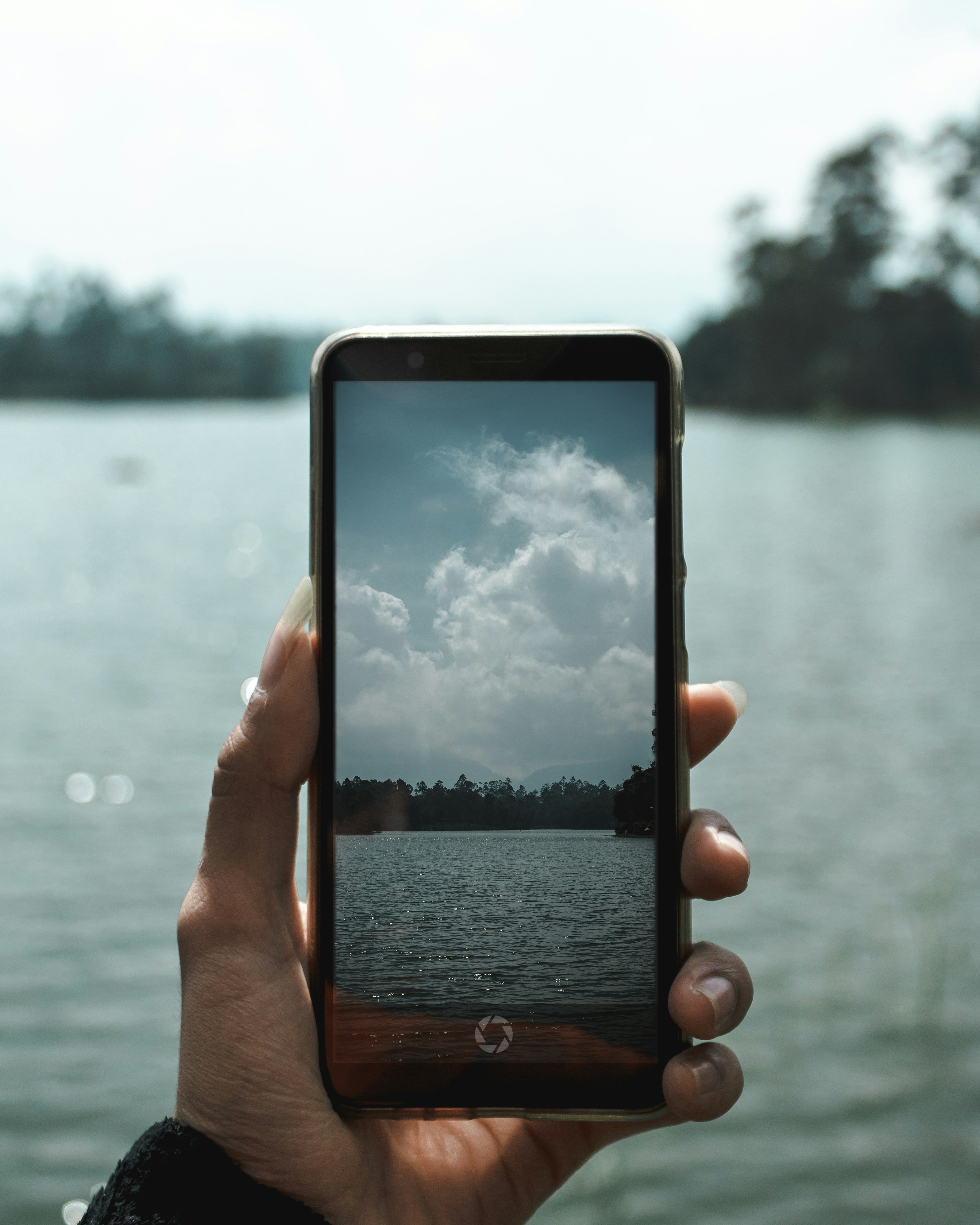 A hand holds a smartphone displaying a serene lake and cloudy sky, framed against a blurred natural backdrop. The scene captures the juxtaposition of technology and nature.