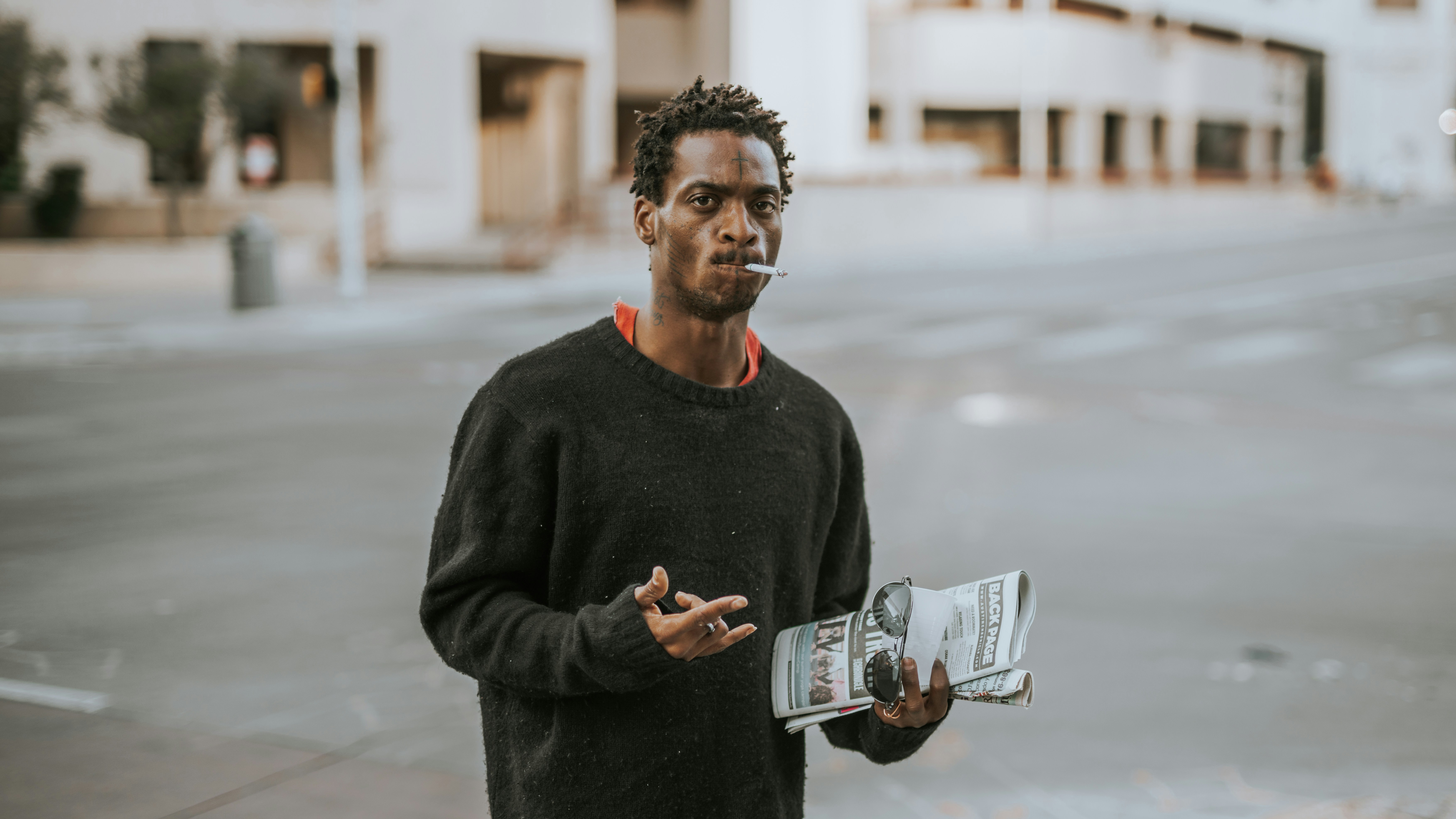 A man stands on a city street, holding a magazine and a cigarette, embodying a moment of introspection amidst urban life.