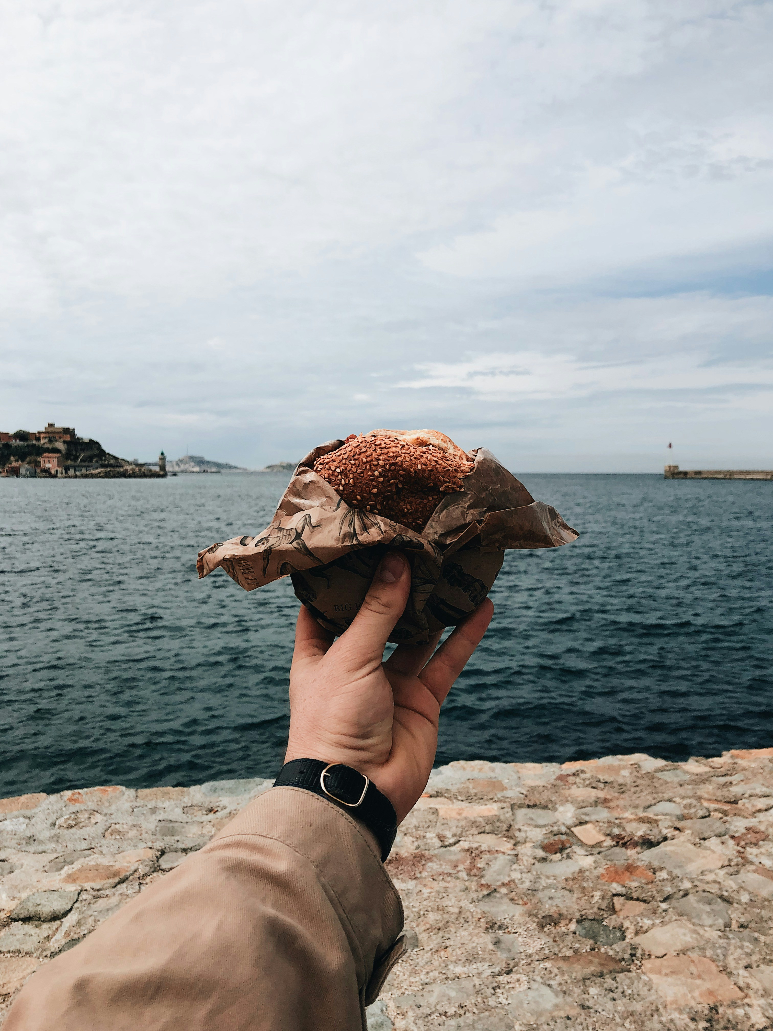 Hand holding a sandwich wrapped in paper against a tranquil seaside backdrop, with distant hills and a lighthouse visible.