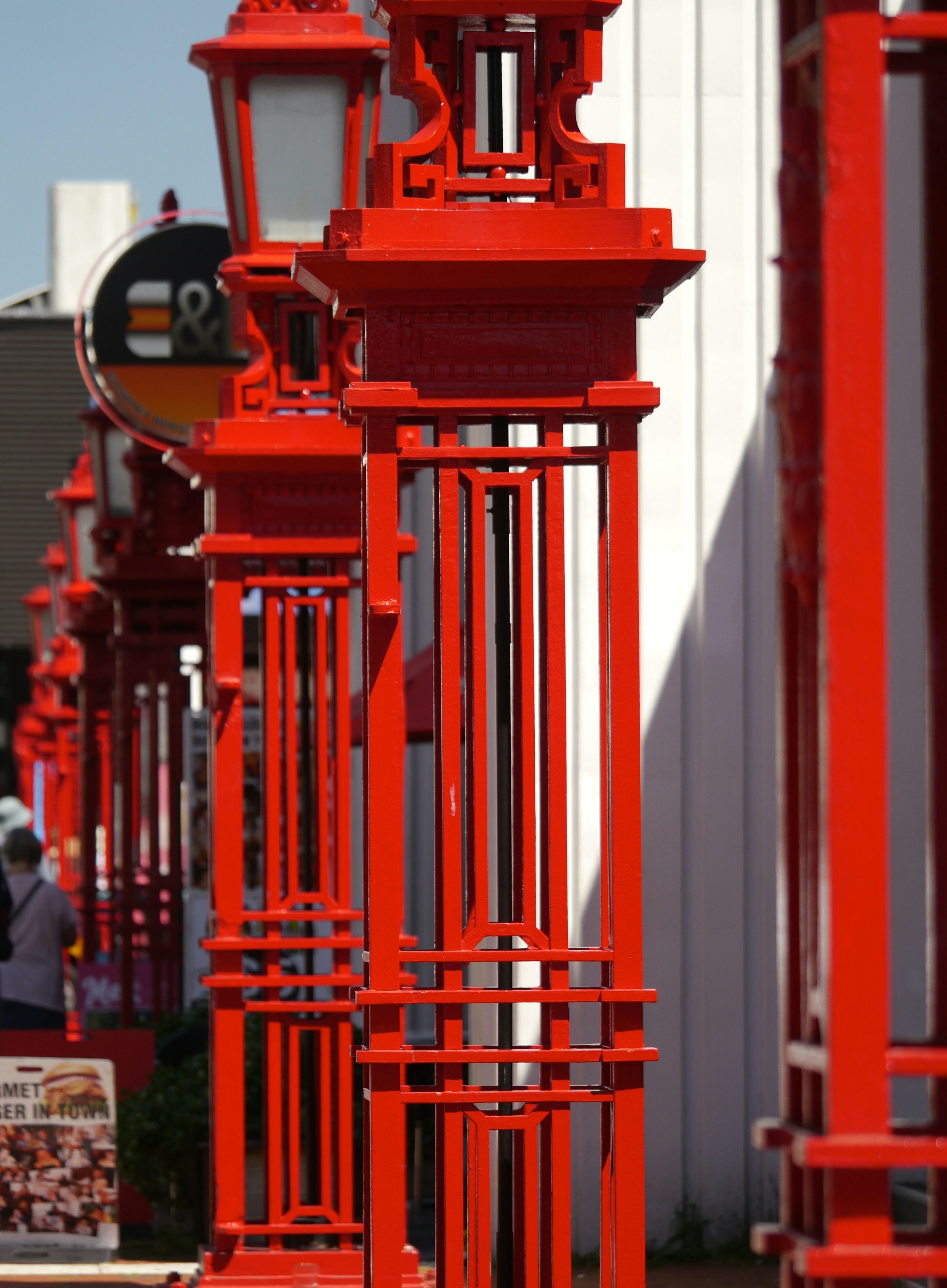 Row of vivid red lantern-style street lamps lines a sunlit urban street. The white facade to the right creates a strong geometric contrast.