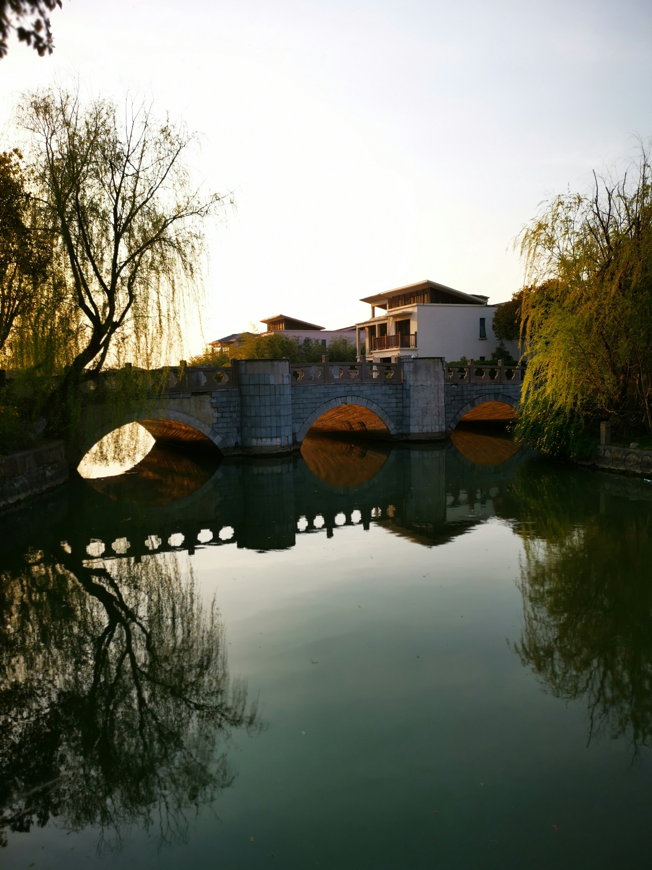 Stone bridge with three arches spans a calm river, its warm brickwork mirrored in the water.