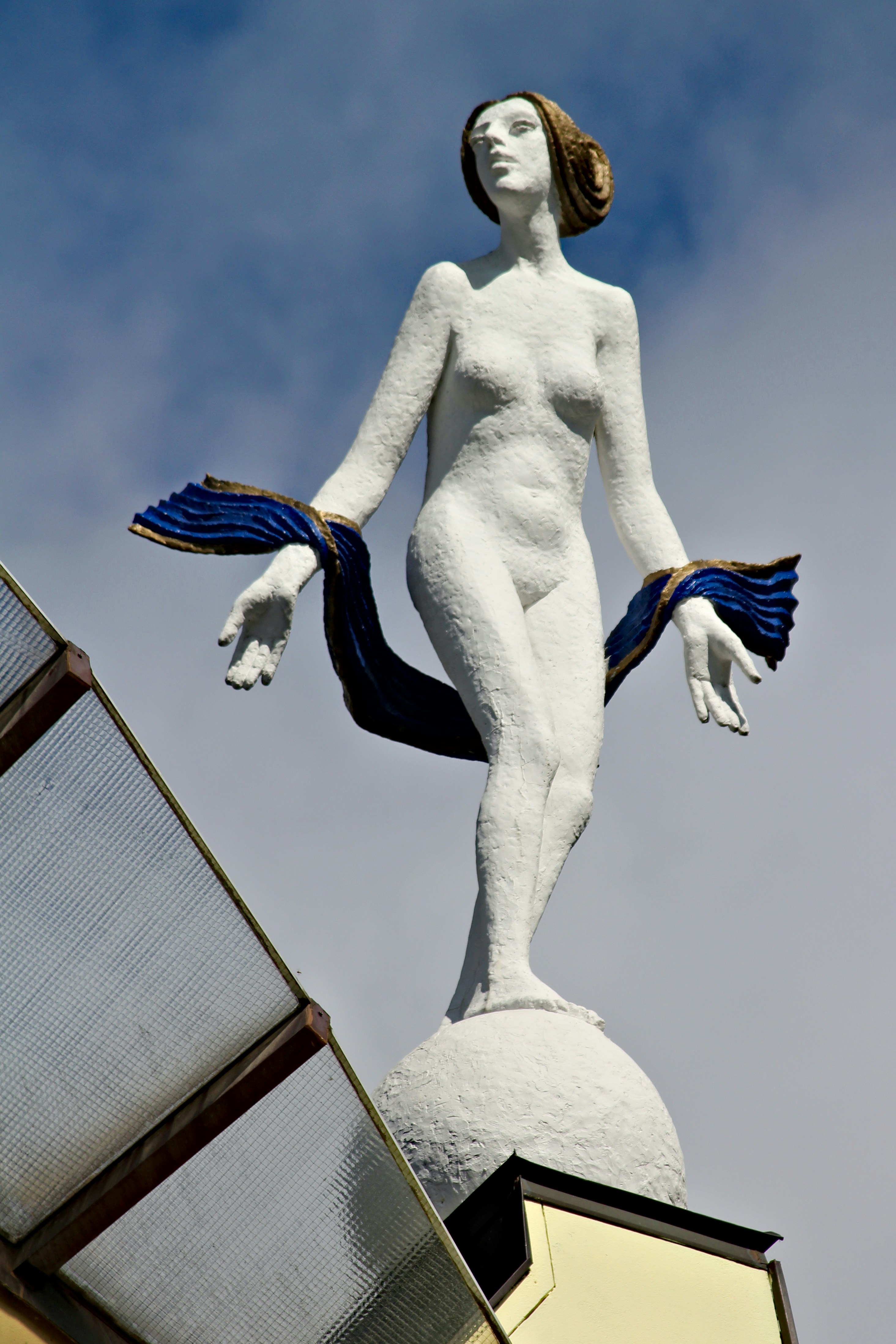 A white sculpture of a female figure in a dynamic pose, adorned with flowing blue ribbons, stands atop a building against a cloudy sky.