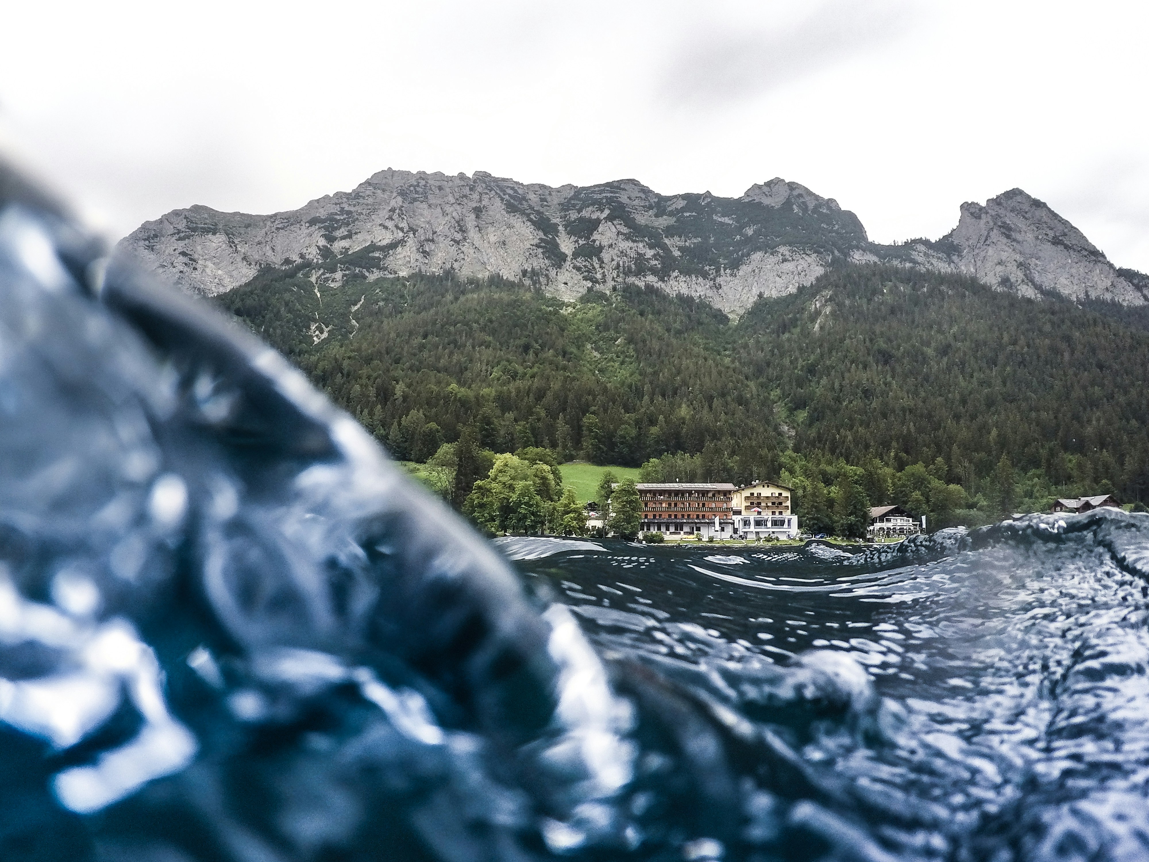 Waves lap against the shoreline, revealing a quaint hotel nestled among lush greenery and towering mountains in the background.
