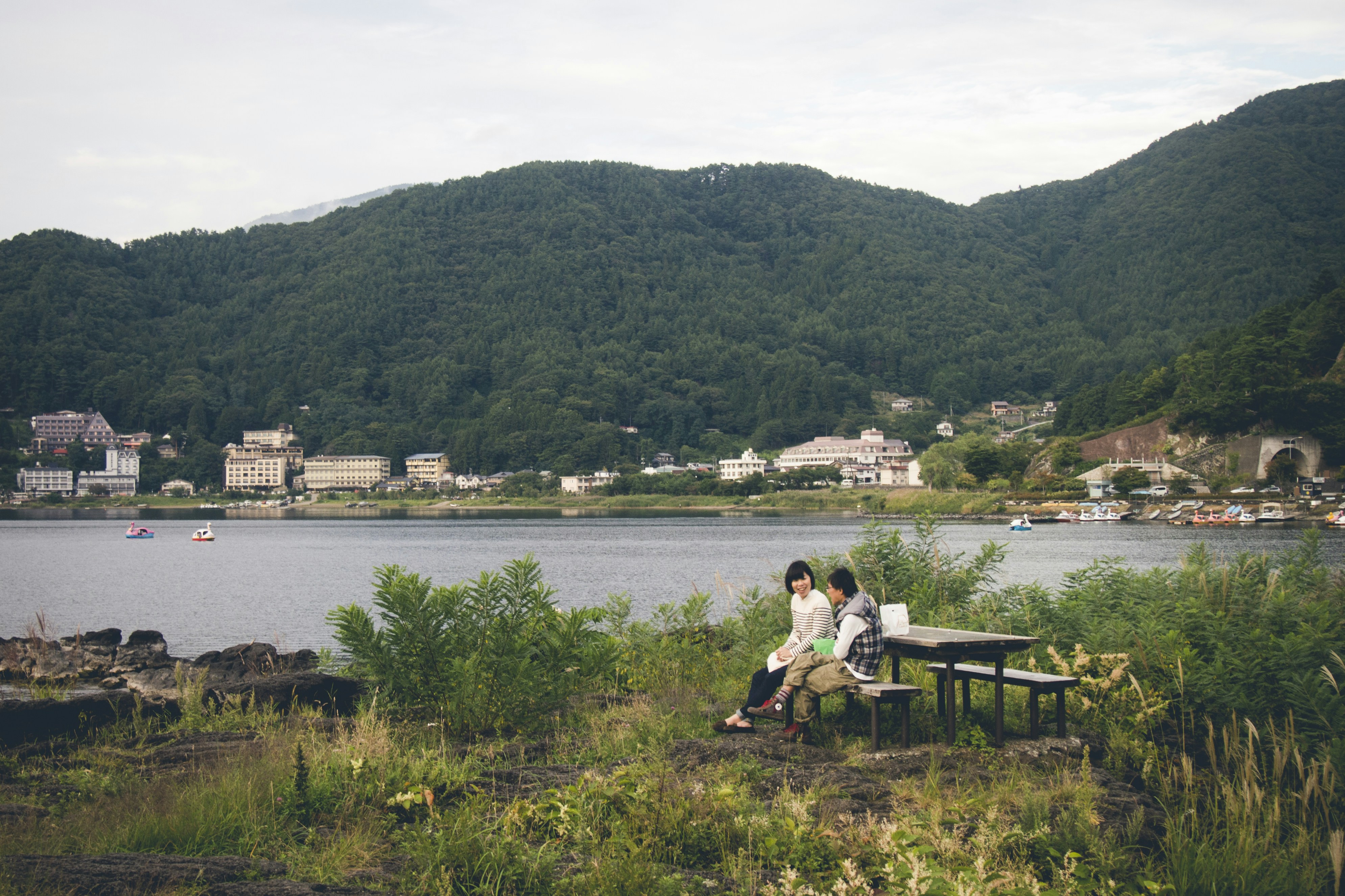 Two people sit on a wooden bench by a calm river, with a small town across the water and forested hills beyond. The scene captures a quiet daytime moment in the countryside.