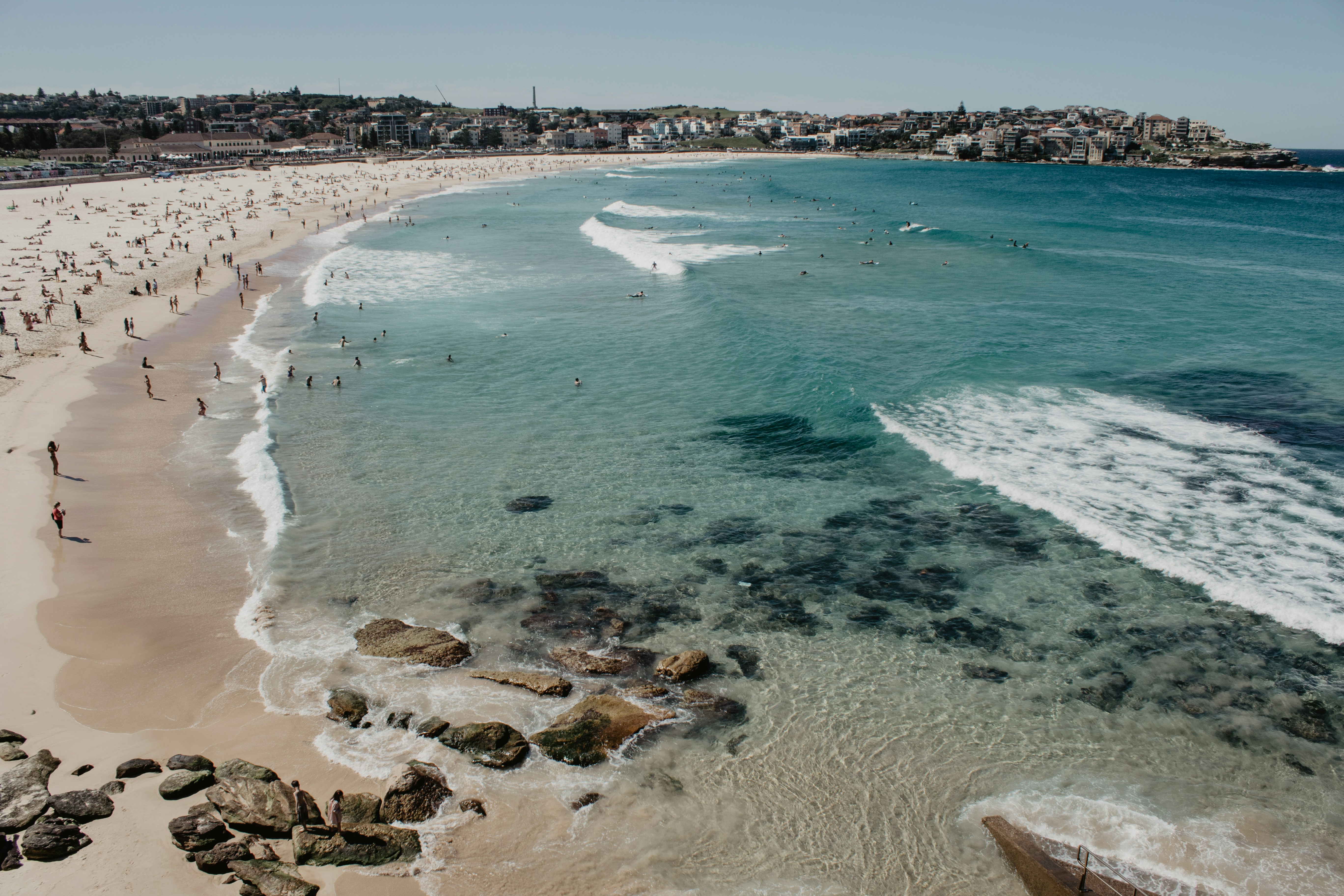 Crowded beach with swimmers in turquoise water, rocky foreground, and distant cityscape.