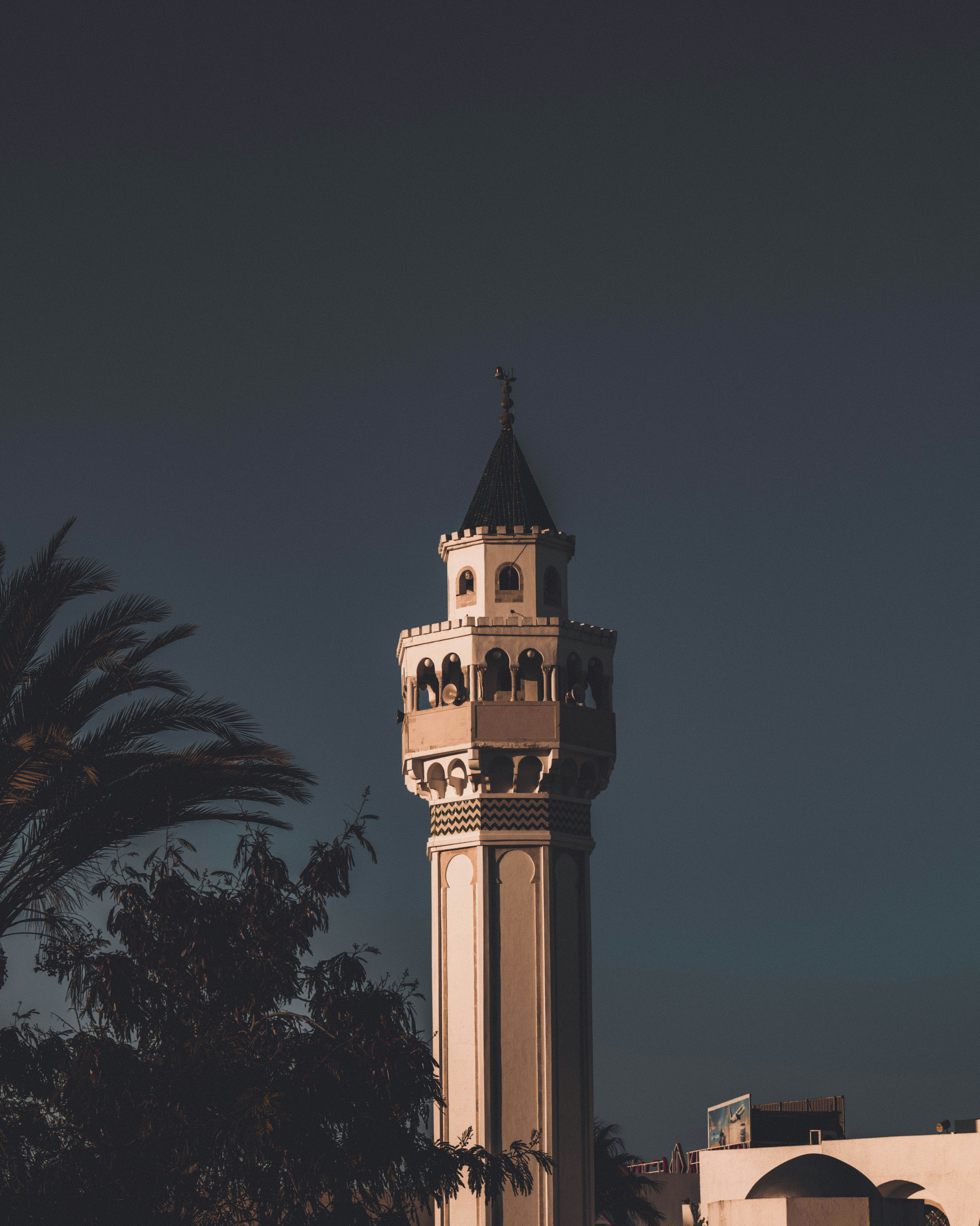 A tall minaret stands against a twilight sky, framed by palm trees, symbolizing a blend of architectural beauty and cultural significance.