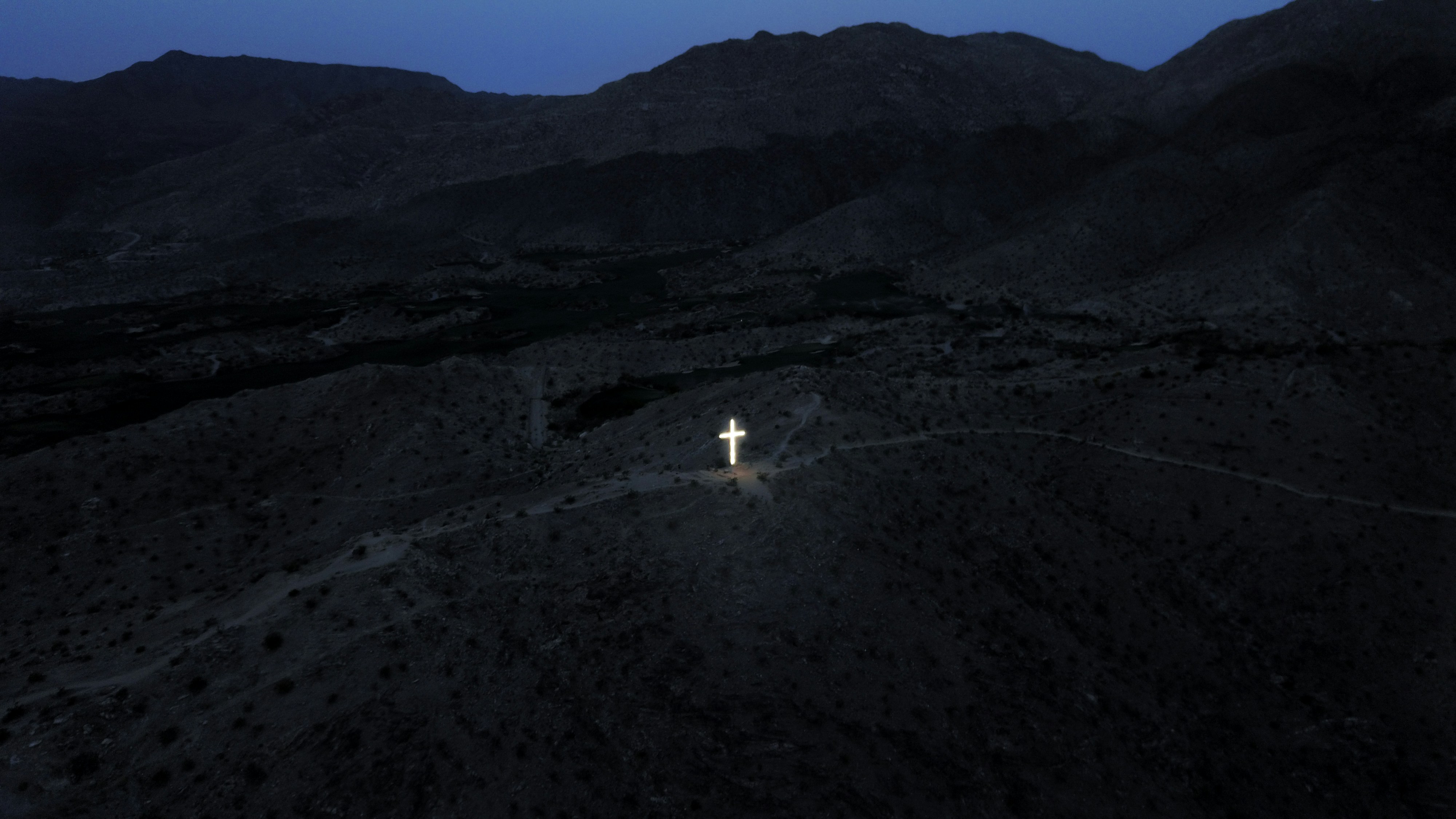A glowing cross stands alone on a mountainside, illuminating the dark landscape under a twilight sky.