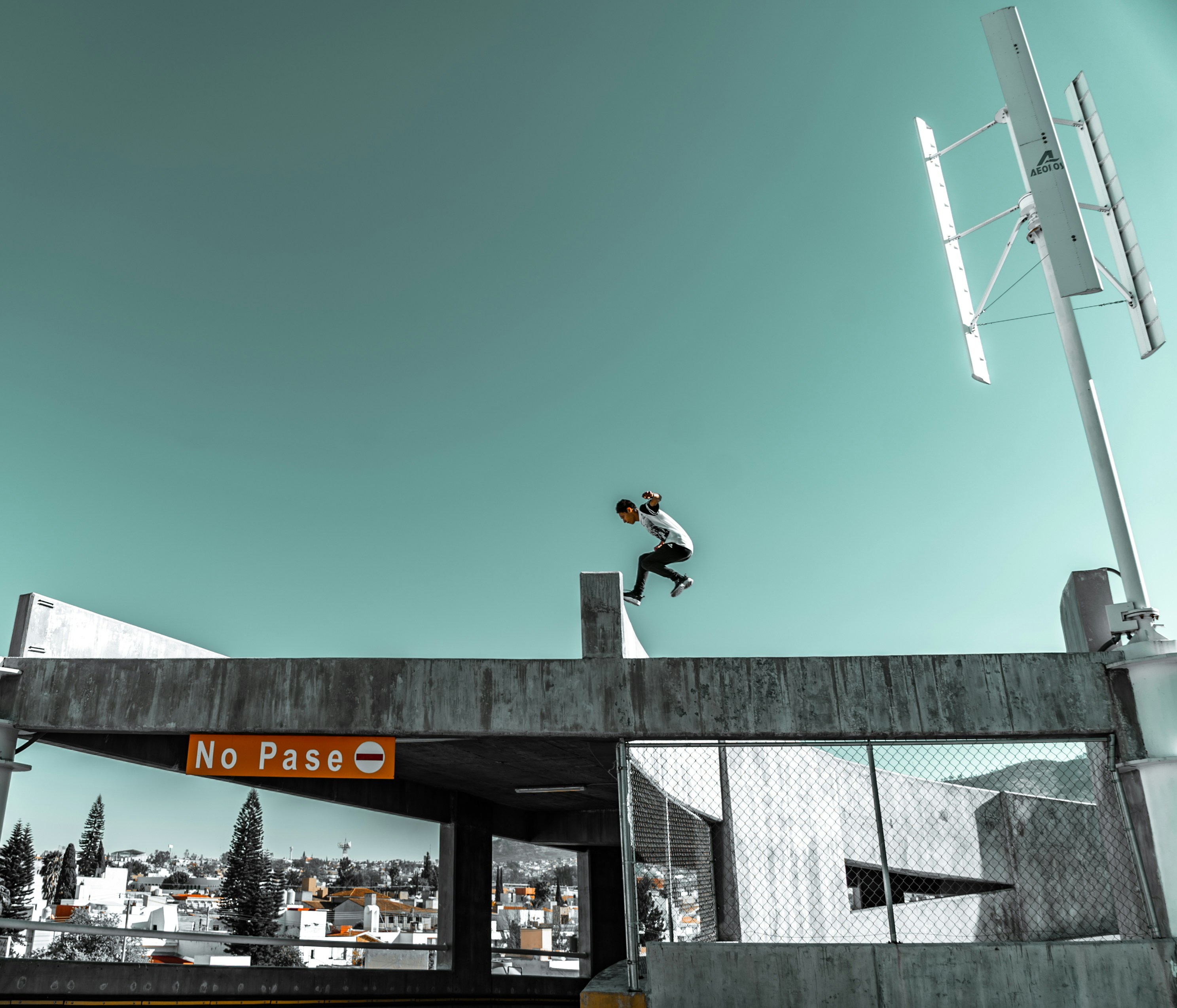 Skateboarder performing a jump off a parking structure with a 'No Pase' sign in the foreground. The urban landscape stretches out below, contrasting with the clear sky.