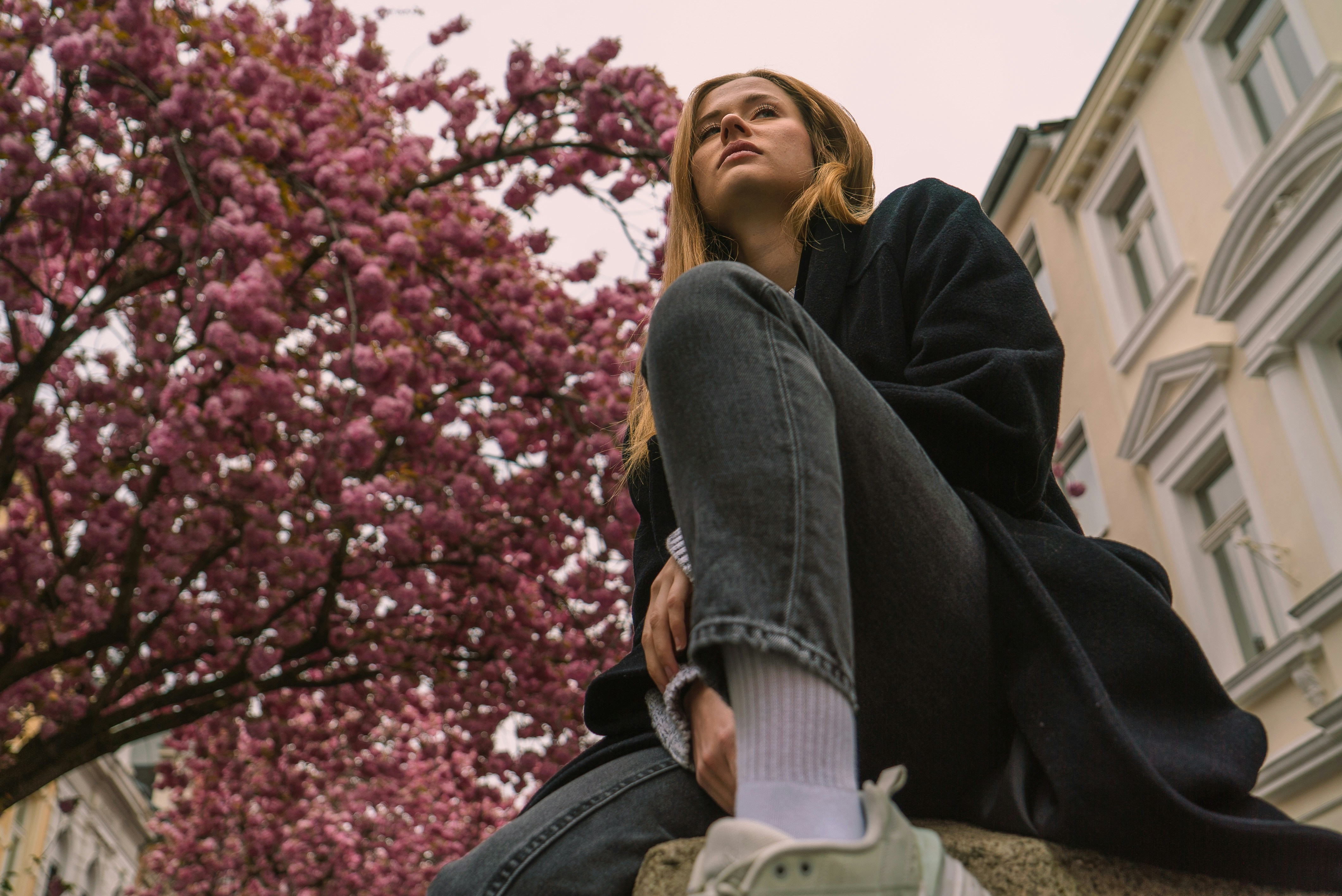 Young woman seated on a stone, gazing thoughtfully, framed by vibrant pink cherry blossoms in full bloom.