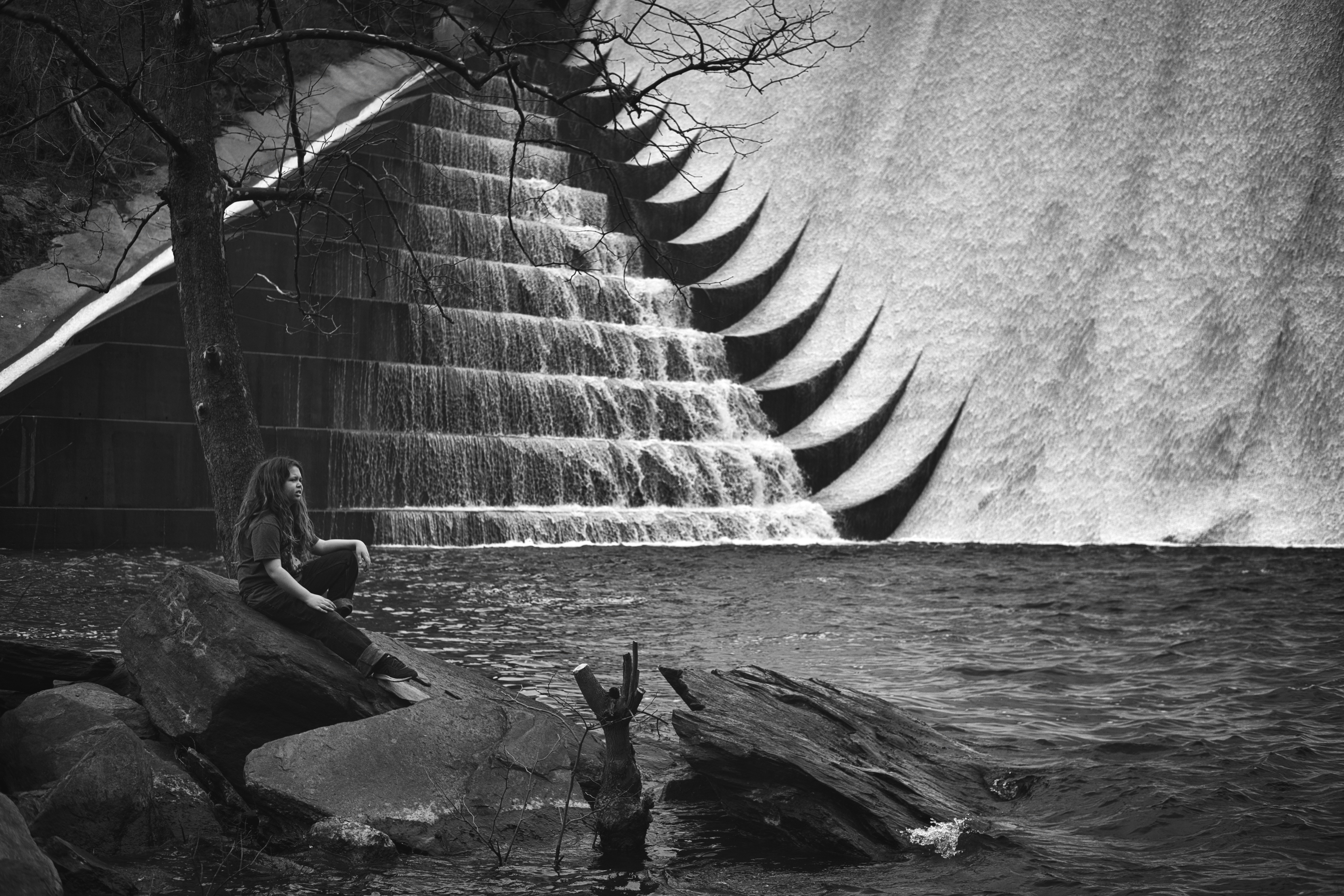 A lone figure sits on a rock by a flowing river, gazing at a dam's cascading water and textured concrete backdrop.