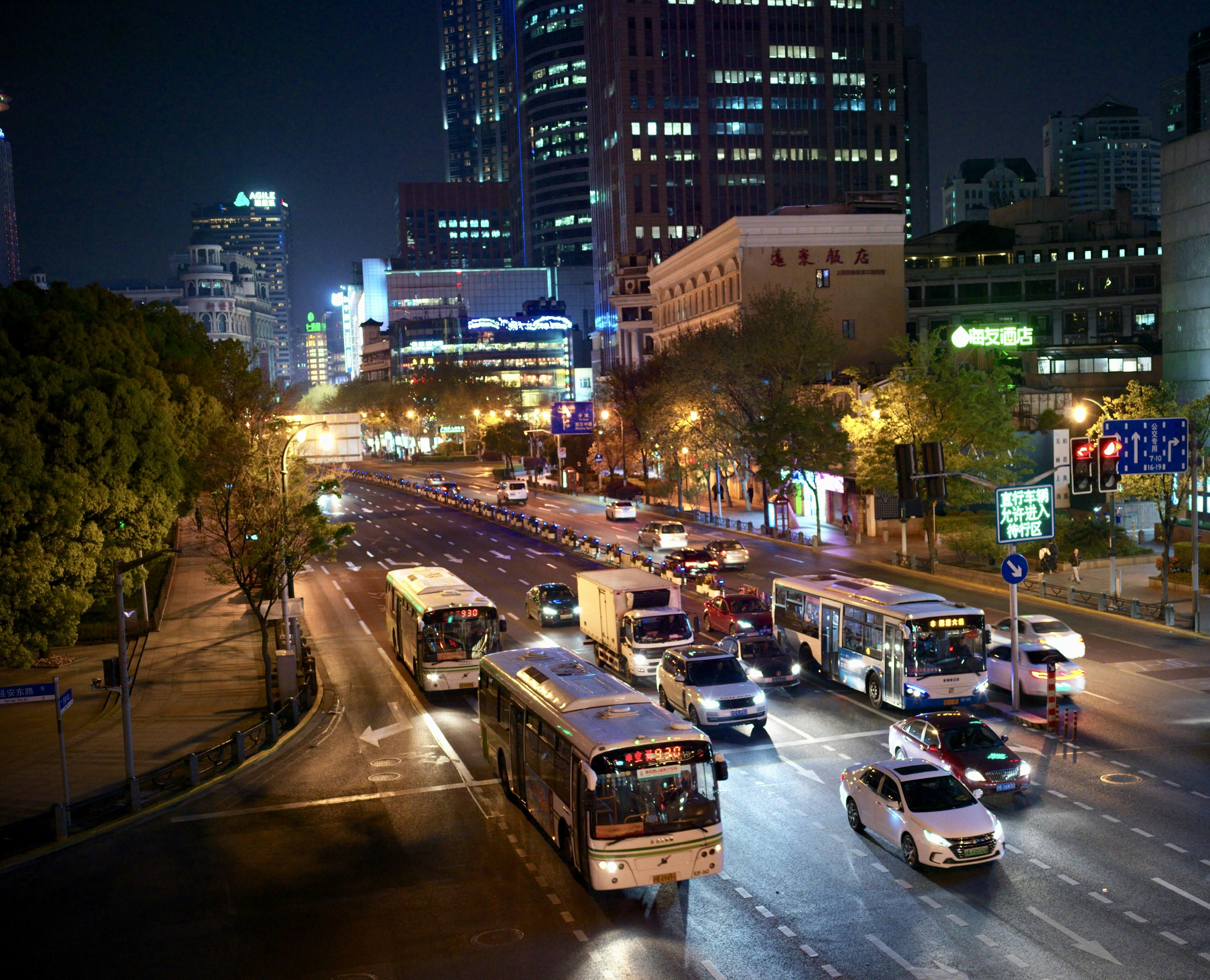 City street bustling with buses and cars under the glow of neon lights at night. The scene captures the vibrant energy of urban life.