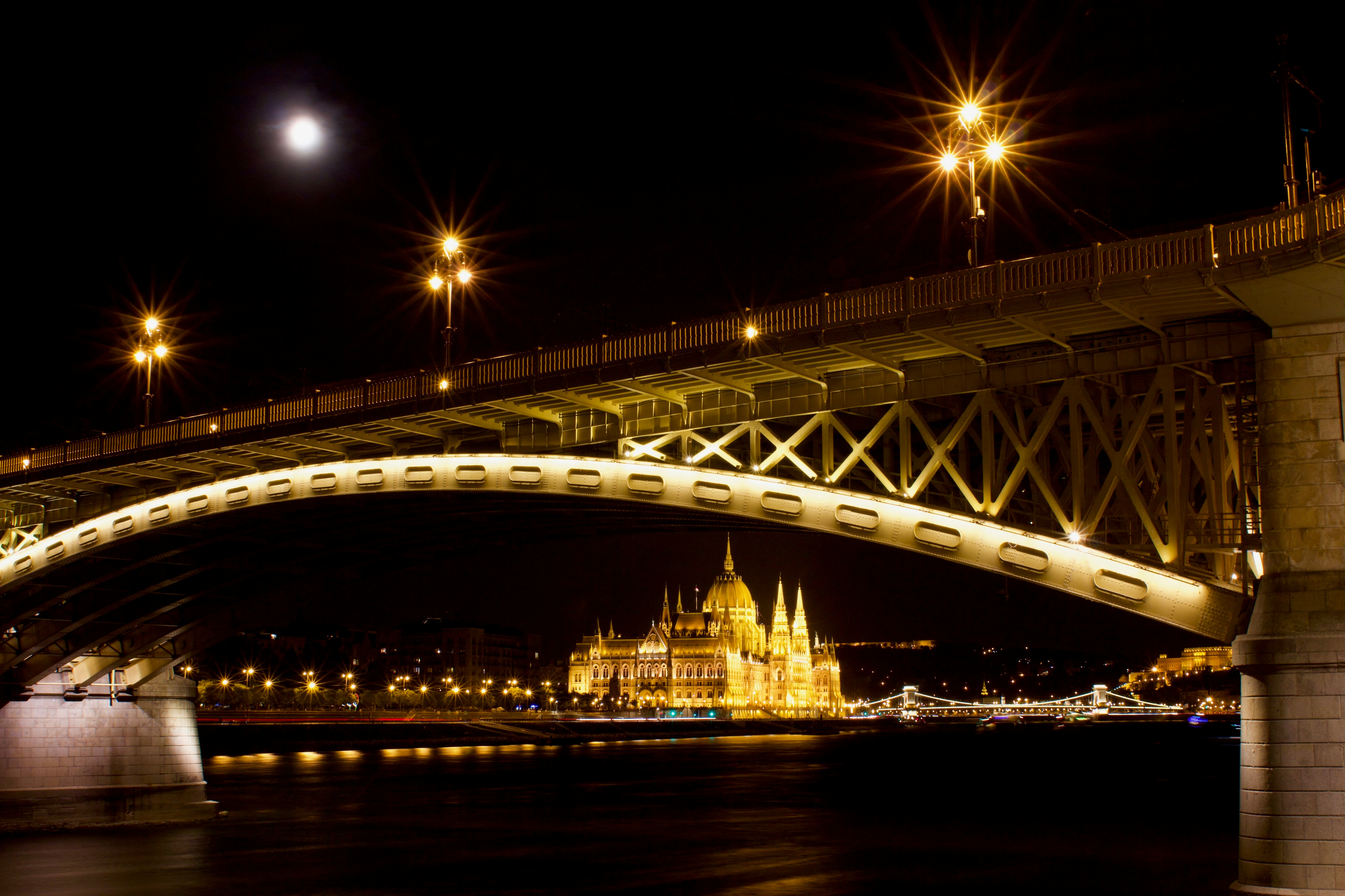 A grand bridge arches over the Danube River, framing the illuminated Hungarian Parliament building against a night sky. The scene captures the essence of Budapest's architectural beauty.