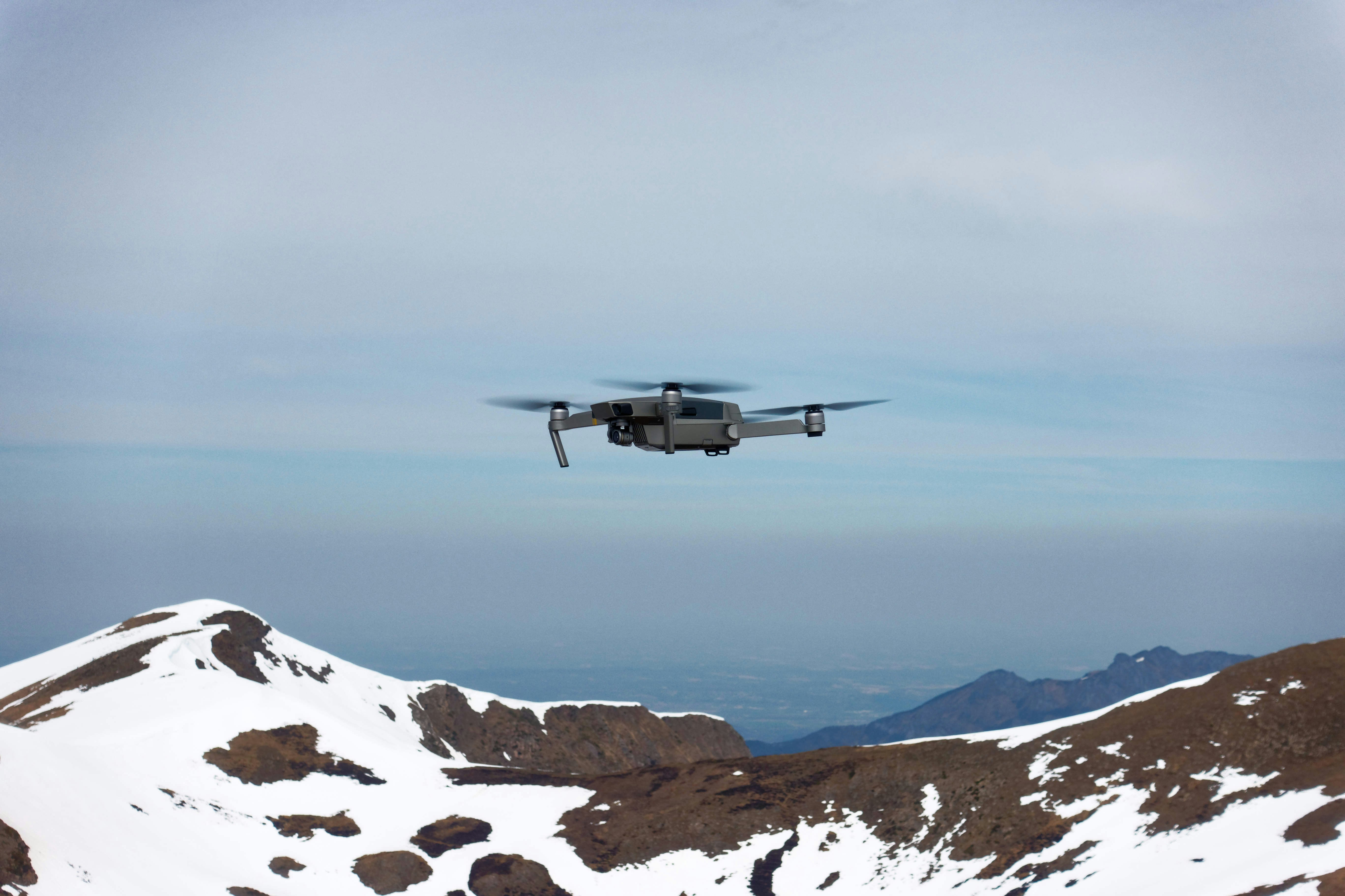 Drone hovering over snow-covered mountains, capturing the vast landscape below. The contrast between the white snow and earthy tones of the terrain is striking.