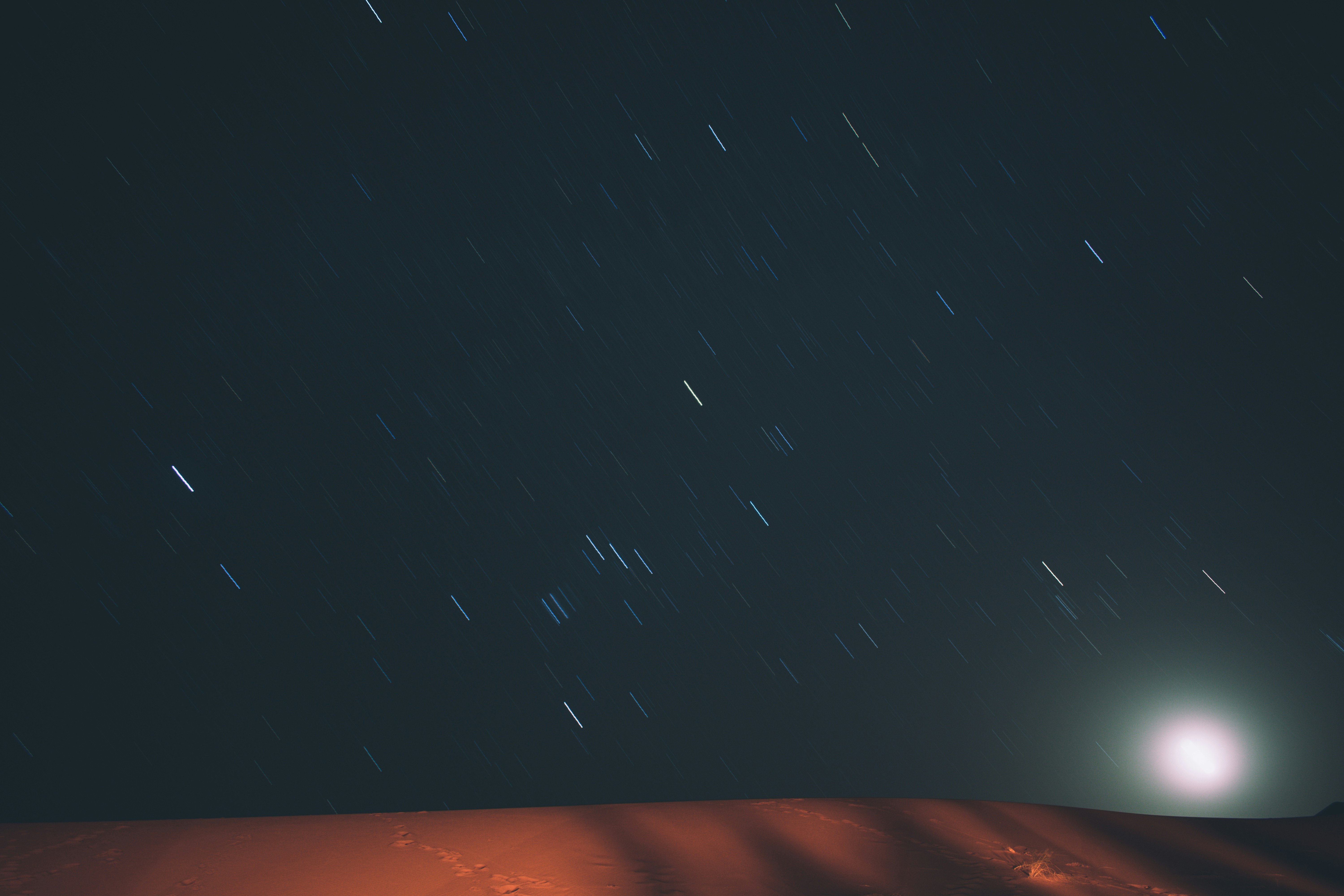 This captivating image captures the serene beauty of a desert under a star-filled sky. The foreground features a gently undulating sand dune bathed in soft, warm light, contrasting with the deep blues and blacks of the night sky. The long exposure creates striking star trails, adding a dynamic element to the tranquil scene, while the moon peeks over the horizon, casting a subtle glow.