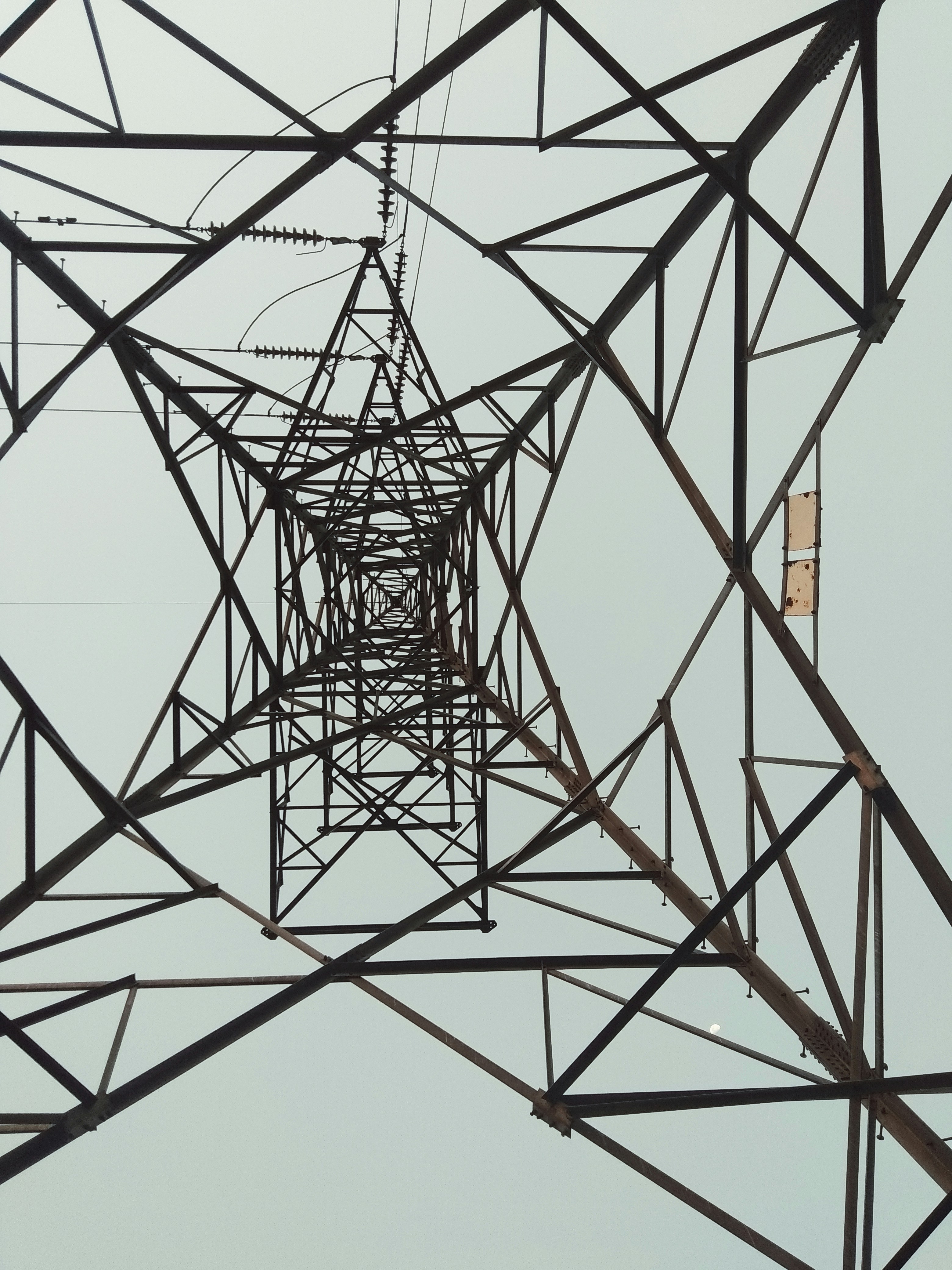 Looking up at a towering electricity pylon, the intricate lattice of steel beams creates a striking geometric pattern against a pale sky.