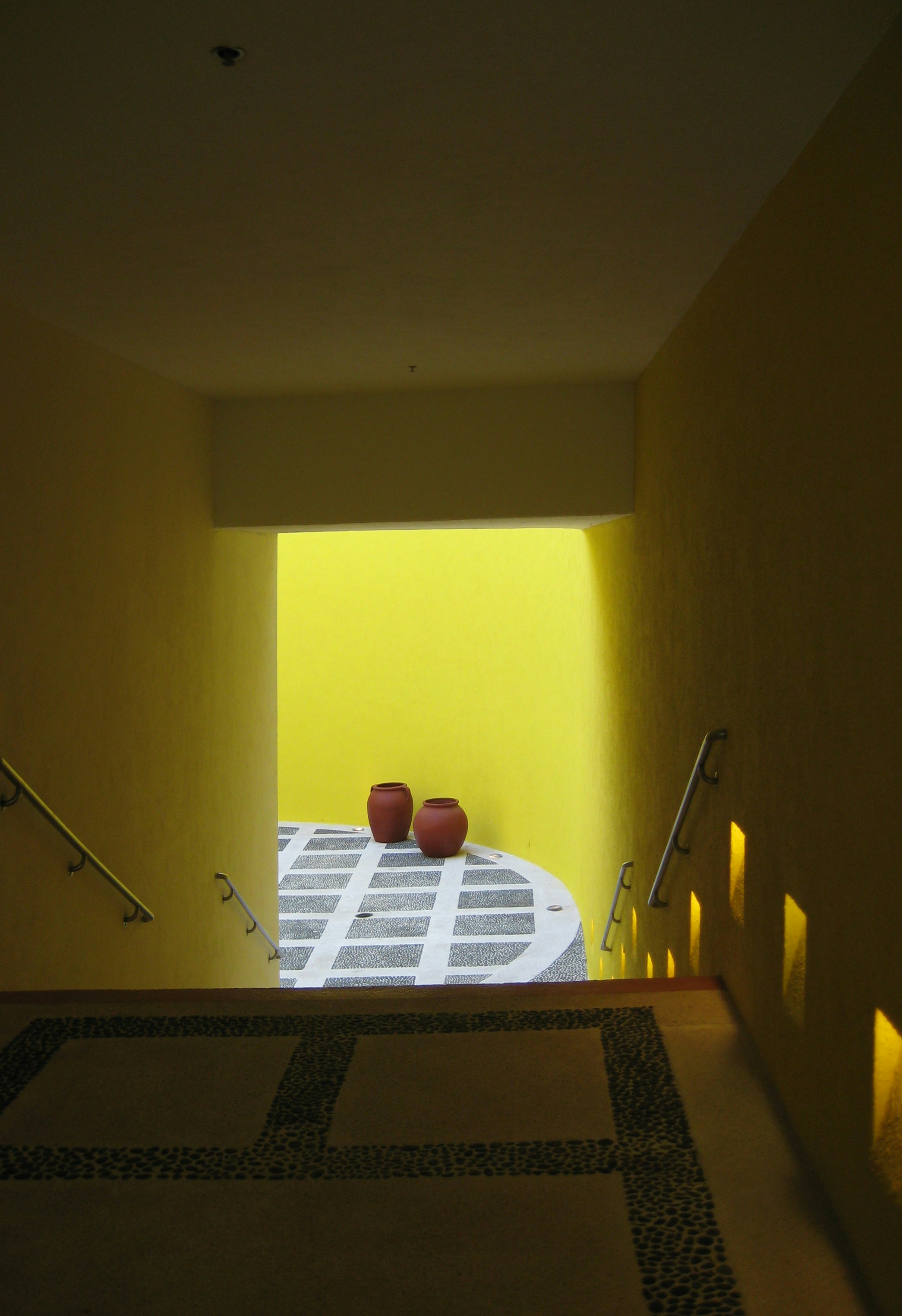 Interior stairwell opens to a sunlit yellow courtyard, with two clay pots at the threshold of a curved, tiled path.