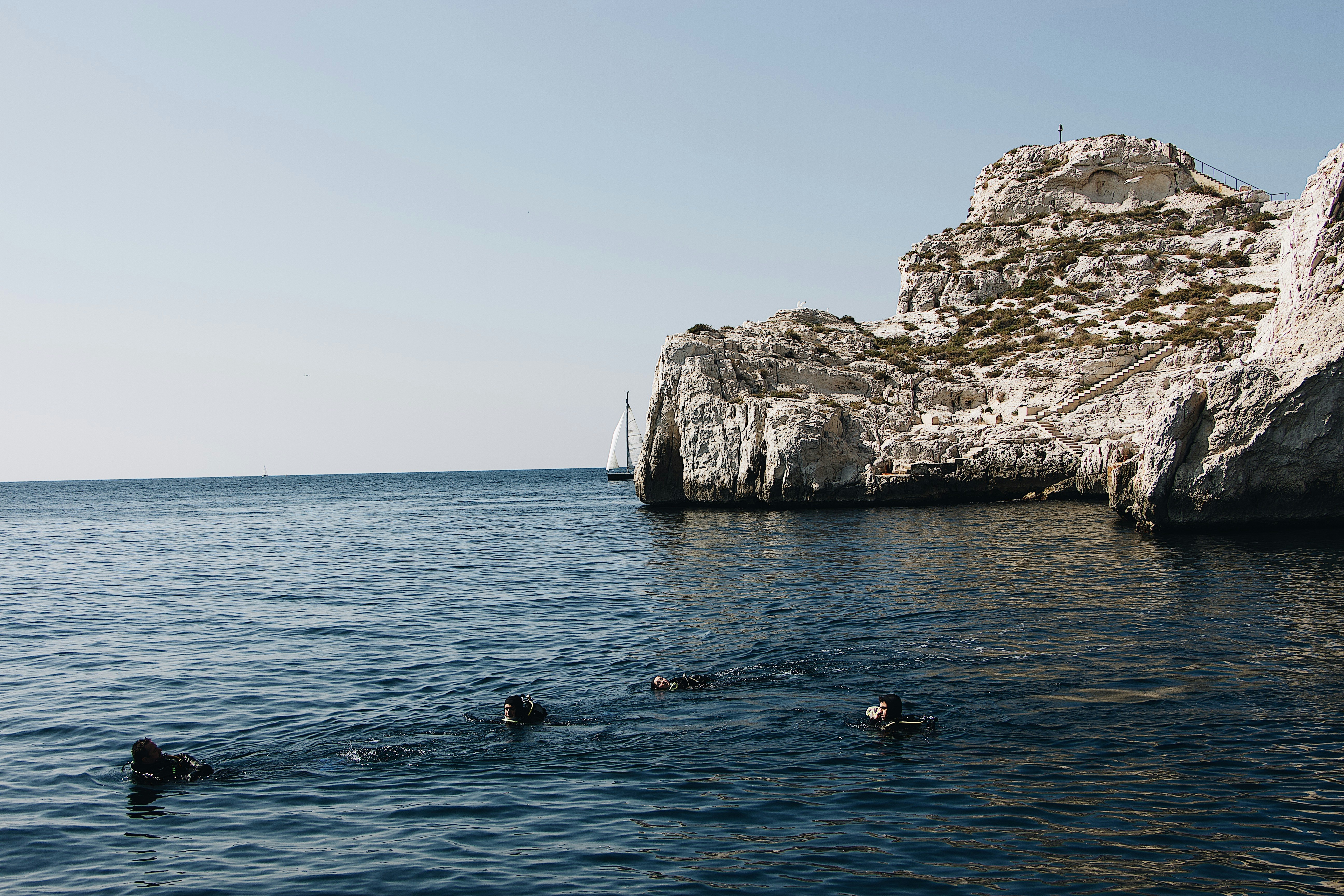 Sailboat glides near rugged cliffs as divers swim in the foreground under a clear sky.