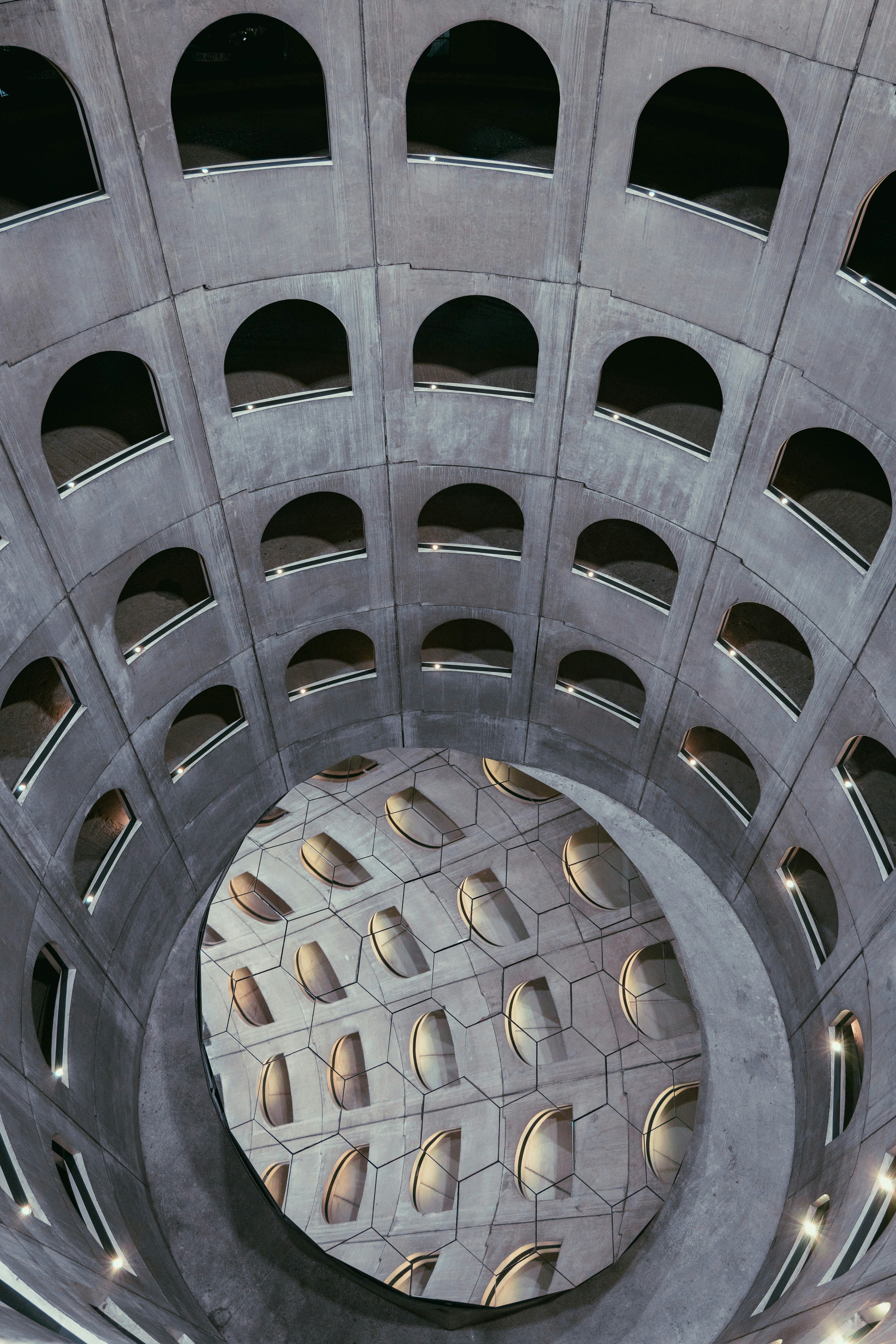 Aerial view of a circular concrete structure showcasing a spiral design with illuminated openings. The interplay of light and shadow creates a dynamic visual effect.