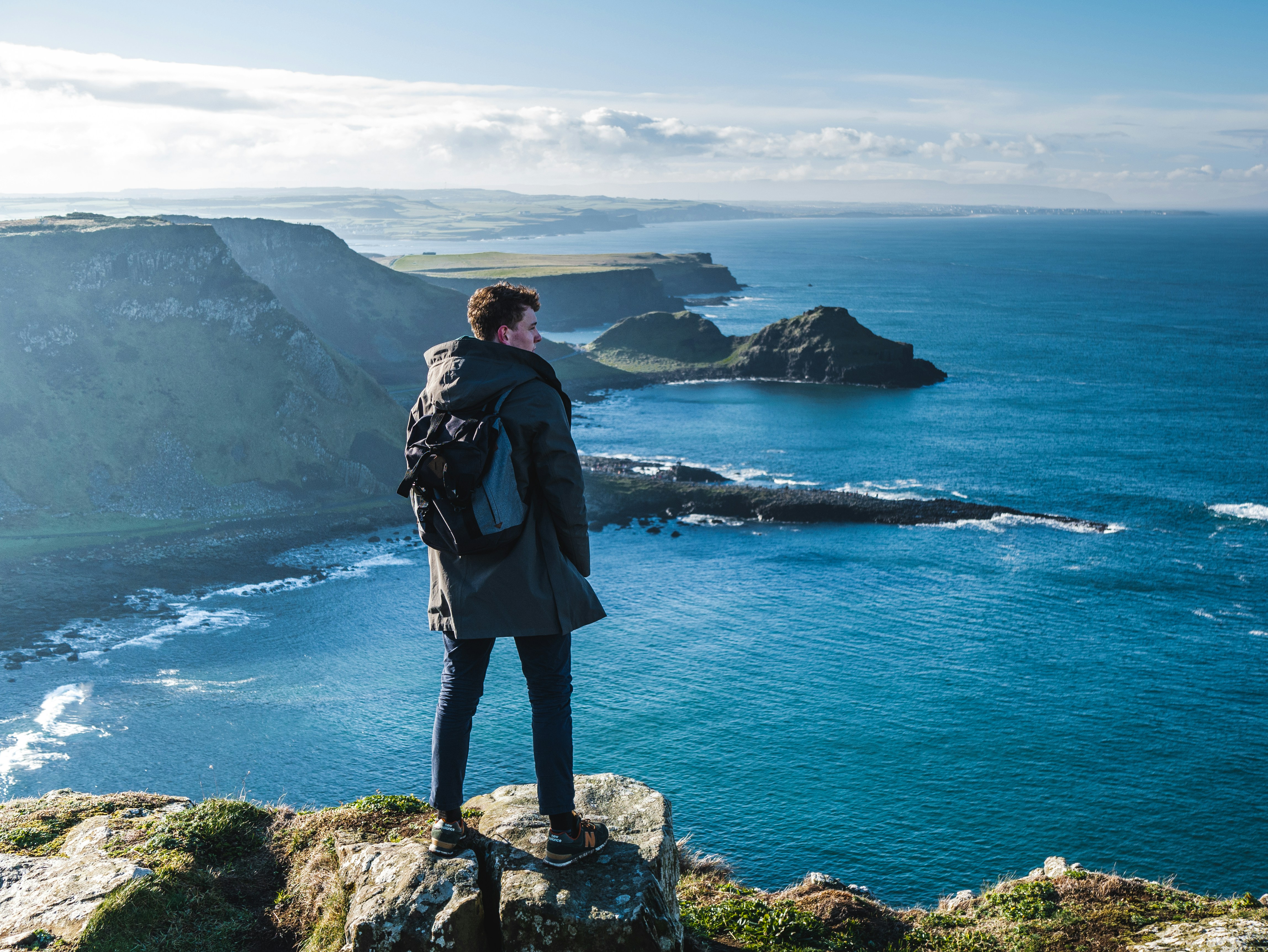 A lone figure stands on a rocky outcrop overlooking a vast coastline, with waves crashing against the cliffs below. The scene captures a moment of solitude and adventure.