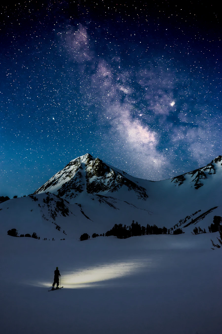 A lone skier stands on a snowy slope beneath a bright Milky Way arc and jagged mountain peaks at night.
