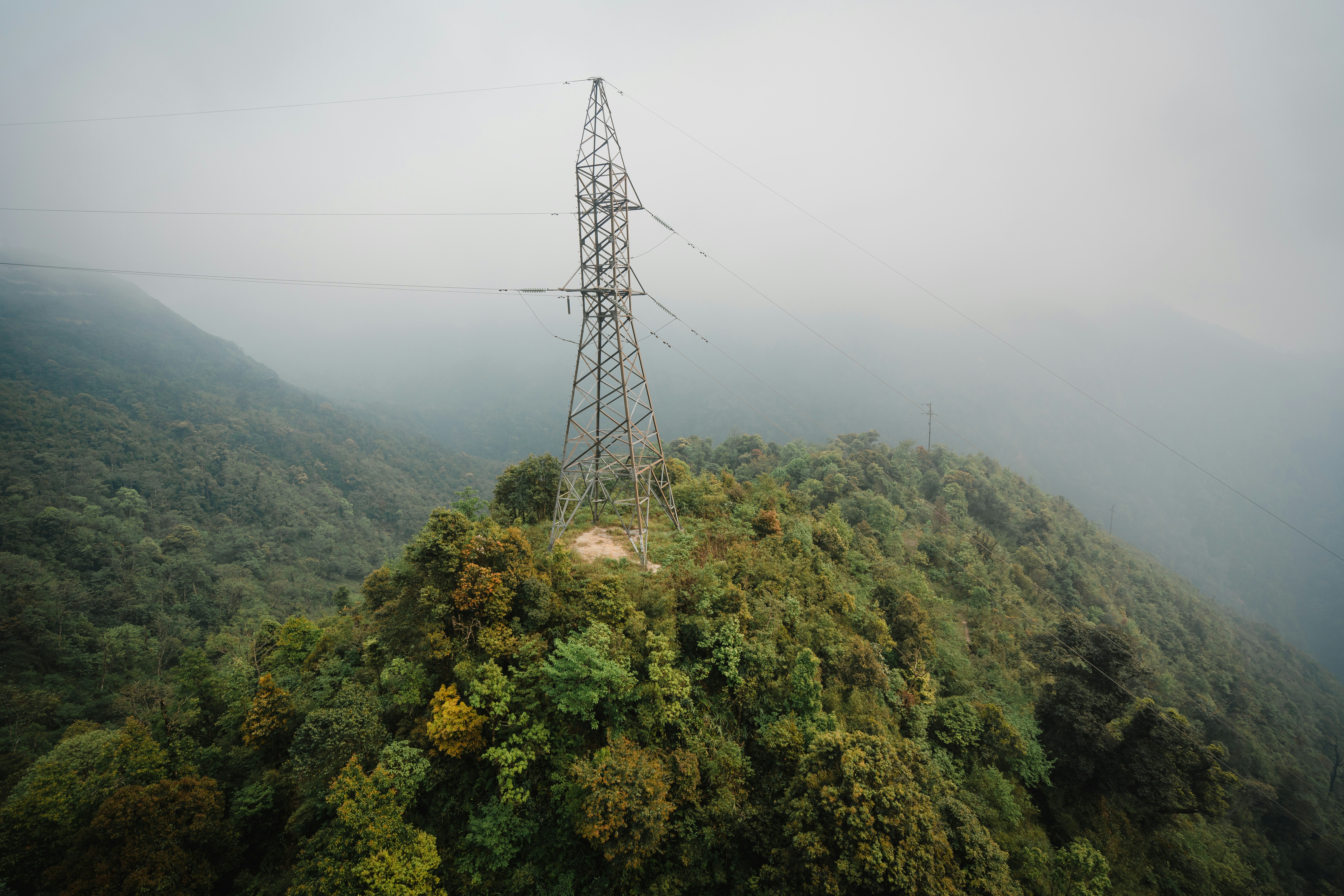 A towering power line structure rises above a lush, green hillside shrouded in mist, symbolizing the intersection of nature and technology.