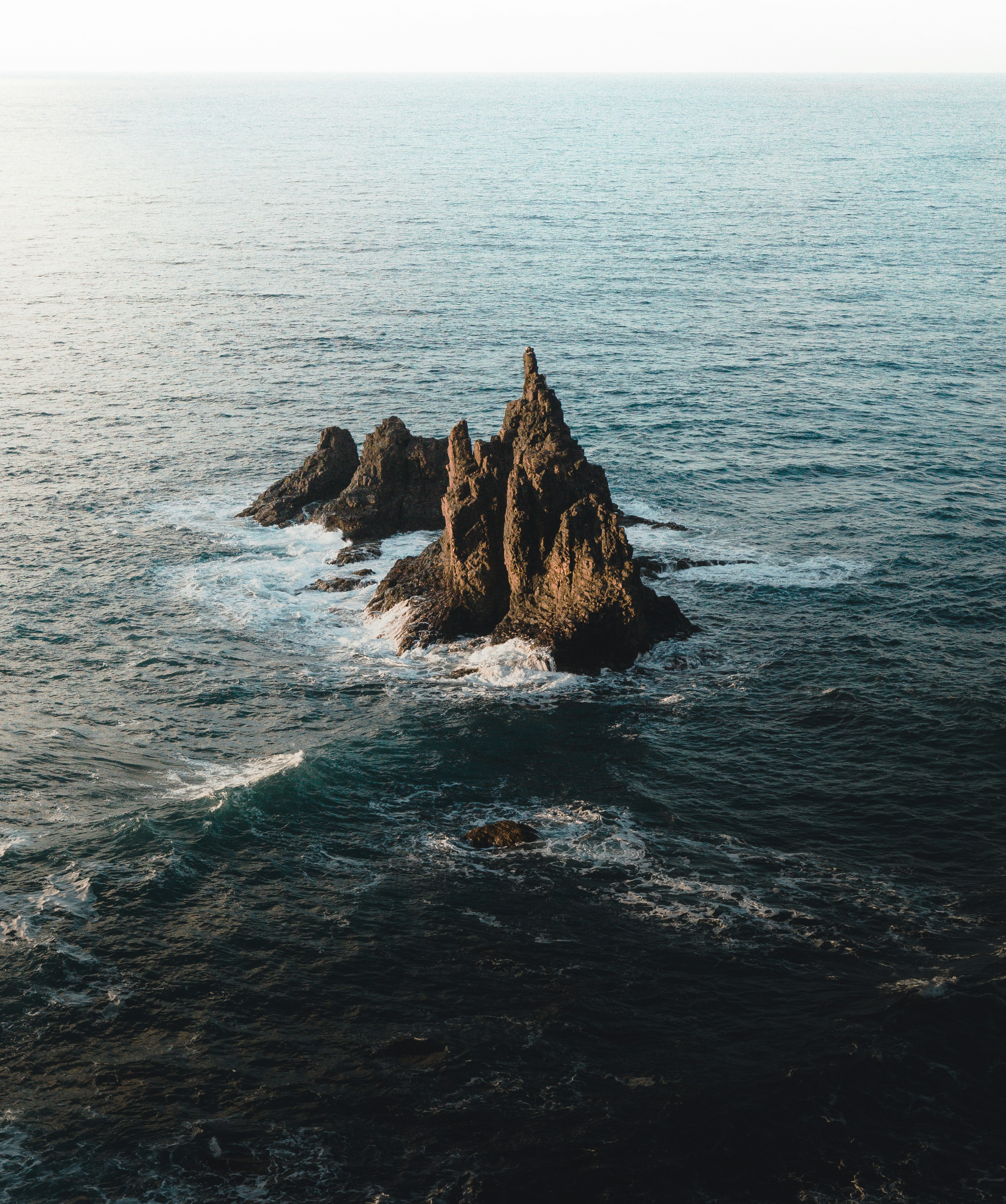 Jagged rock formation surrounded by ocean waves under a soft sunset glow.