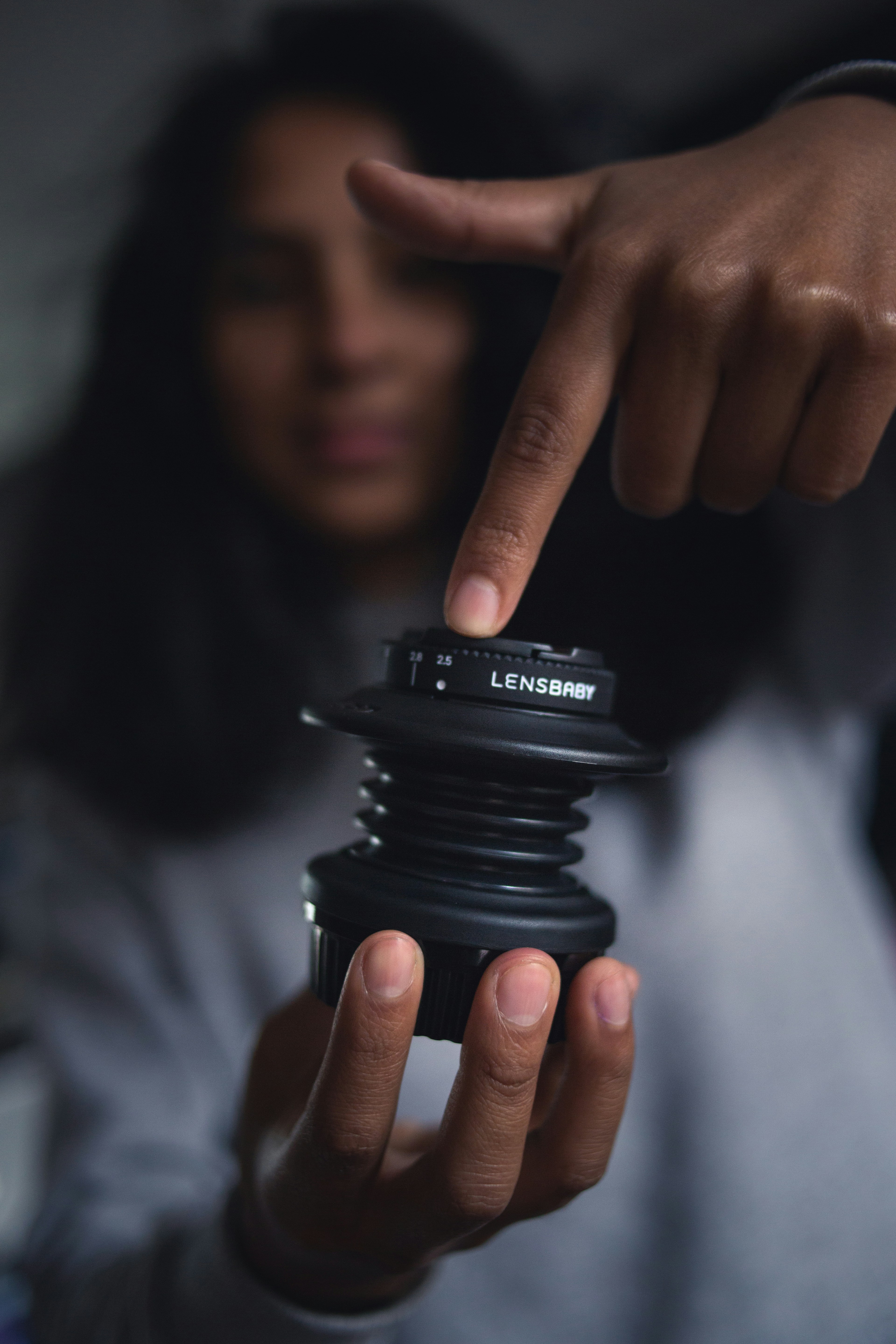 A person points at a Lensbaby lens, showcasing its unique design and functionality. The background is softly blurred, emphasizing the lens in hand.