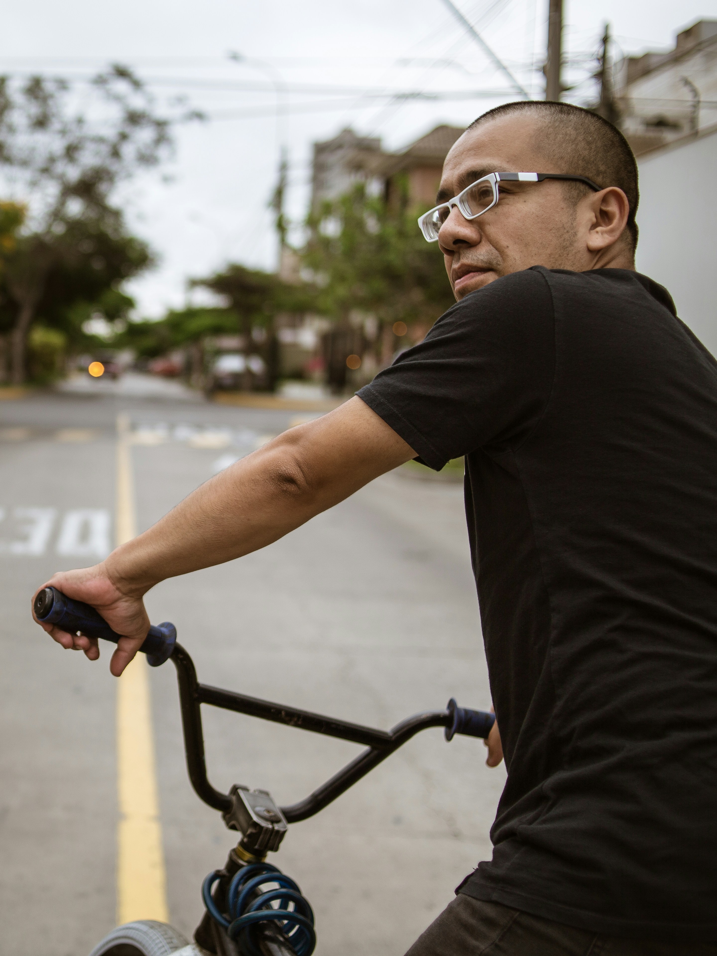 Man in a black shirt riding a bicycle down an urban street, with trees lining the road and buildings in the background.