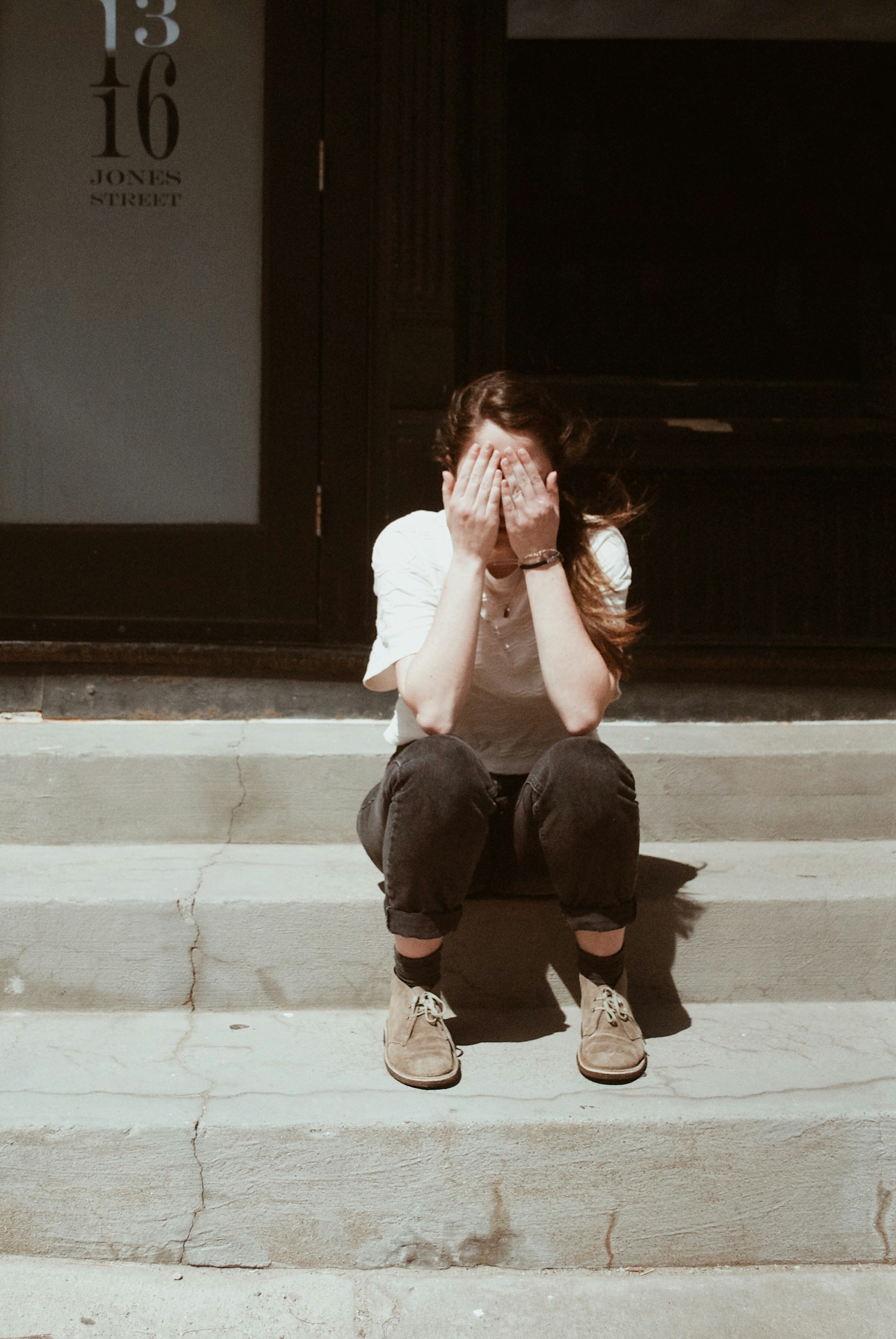 A young woman sits on steps, covering her face with her hands, conveying a moment of vulnerability. The backdrop features a door with a street address, adding context to her surroundings.