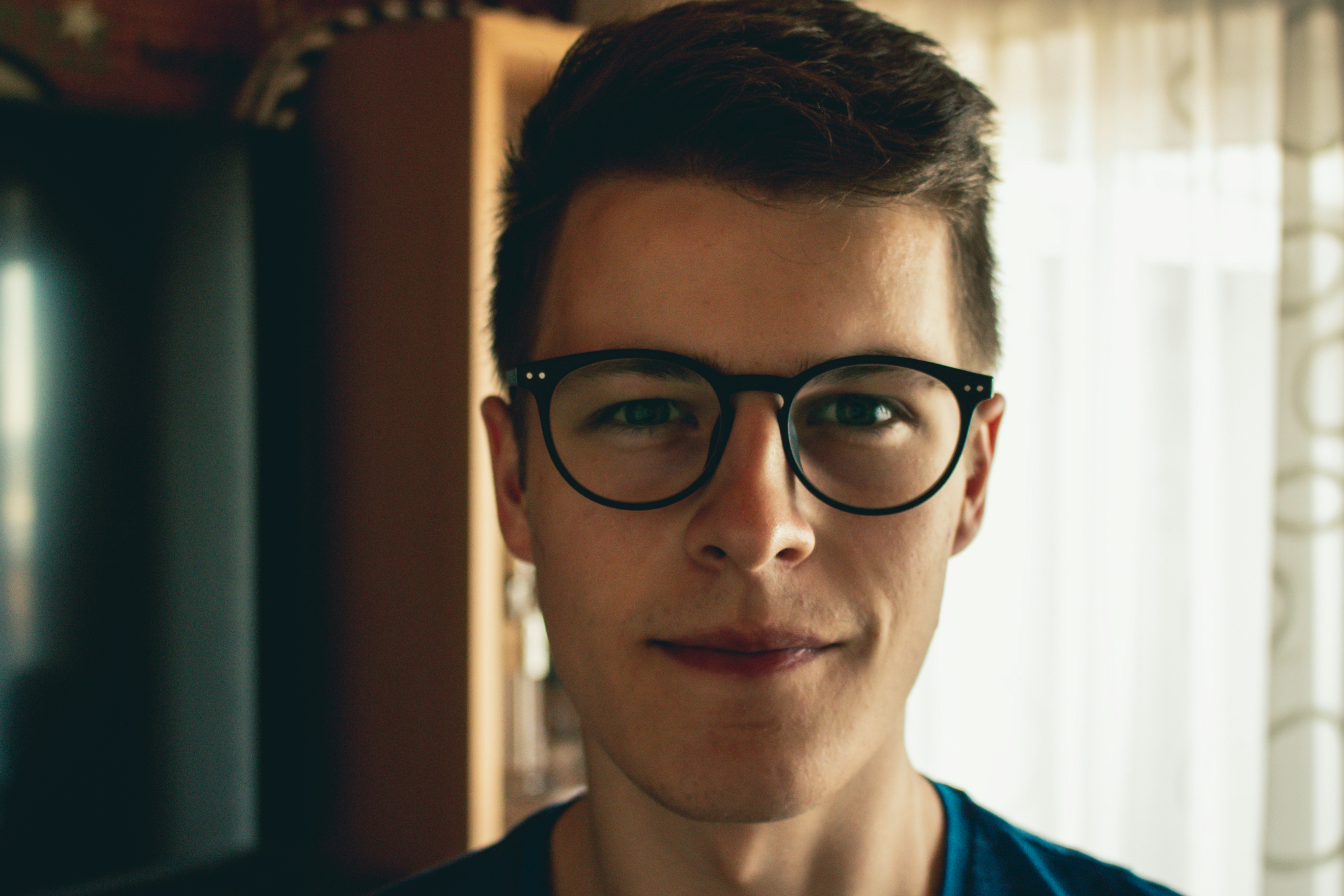 Young man with glasses in a softly lit room, sunlight filtering through curtains.