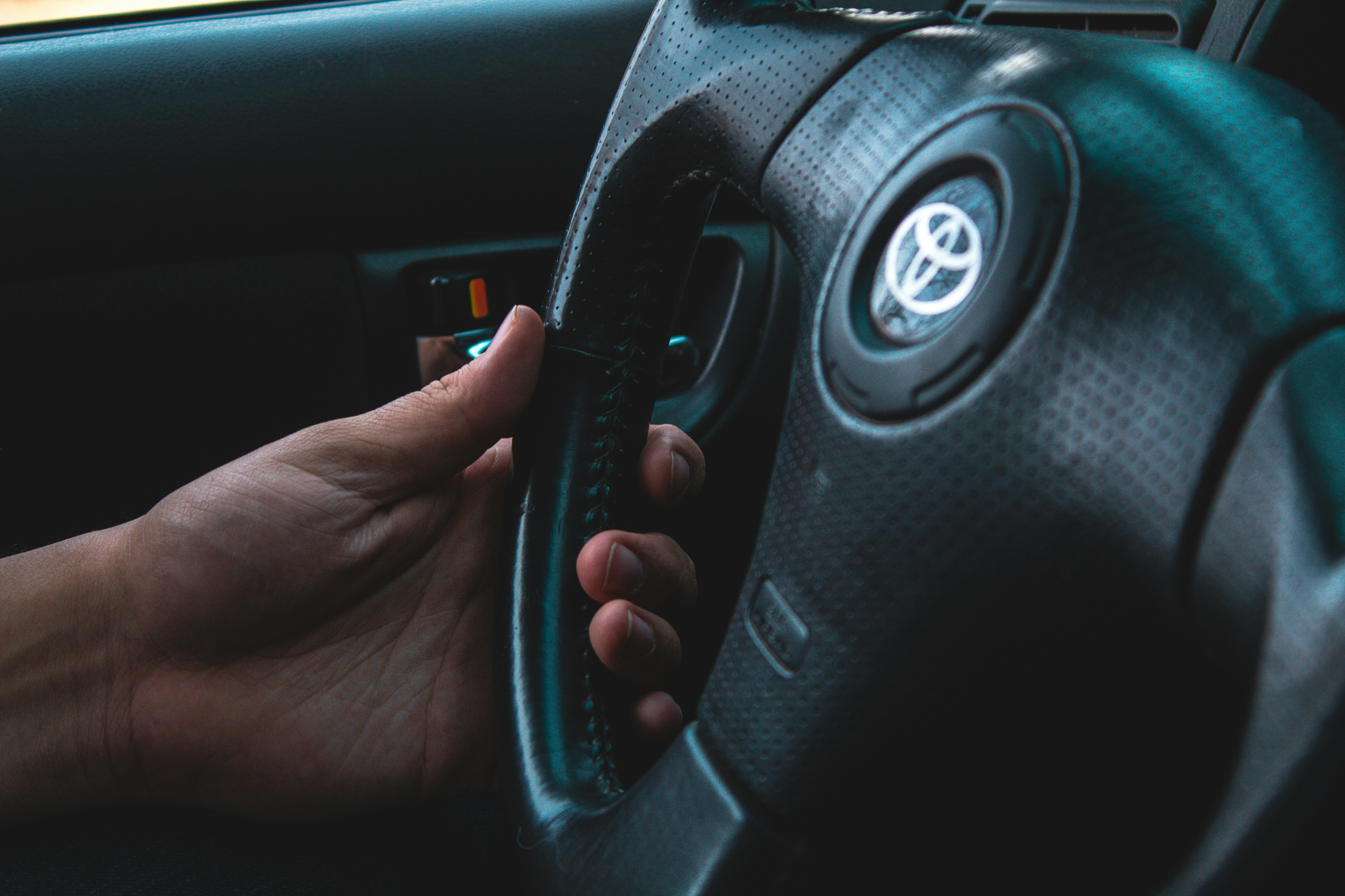 Close-up of a hand on a car steering wheel, highlighting the logo and texture in low light.